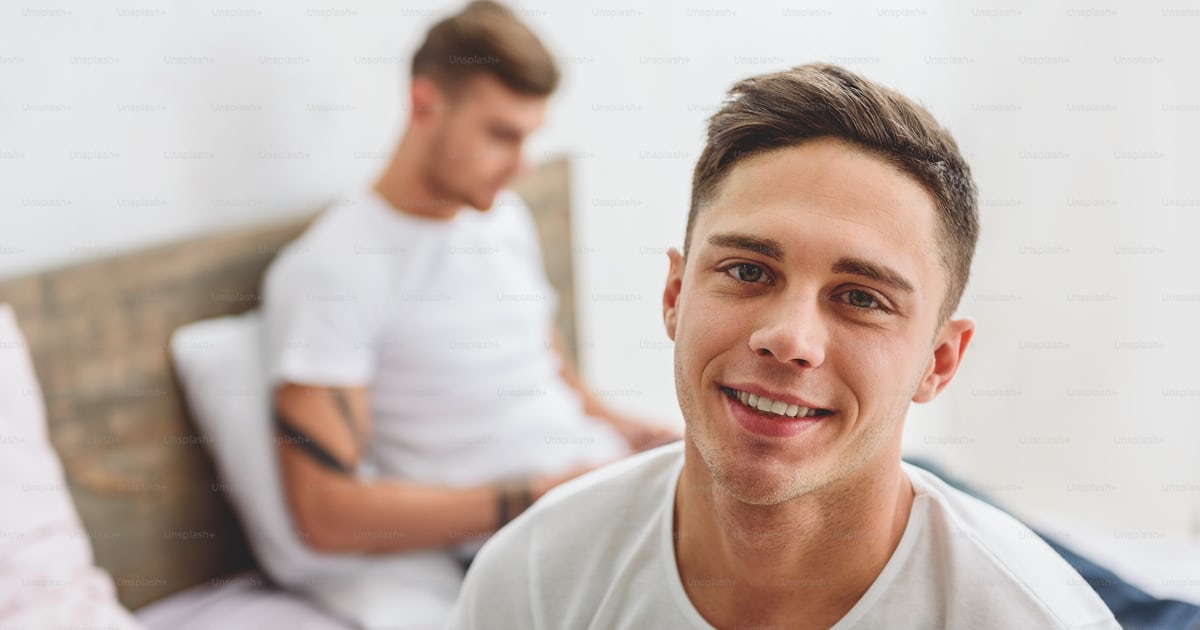 Un Jeune Gay Heureux Regarde La Caméra Et Rit Son Partenaire Est Allongé Sur Le Lit Et Utilise