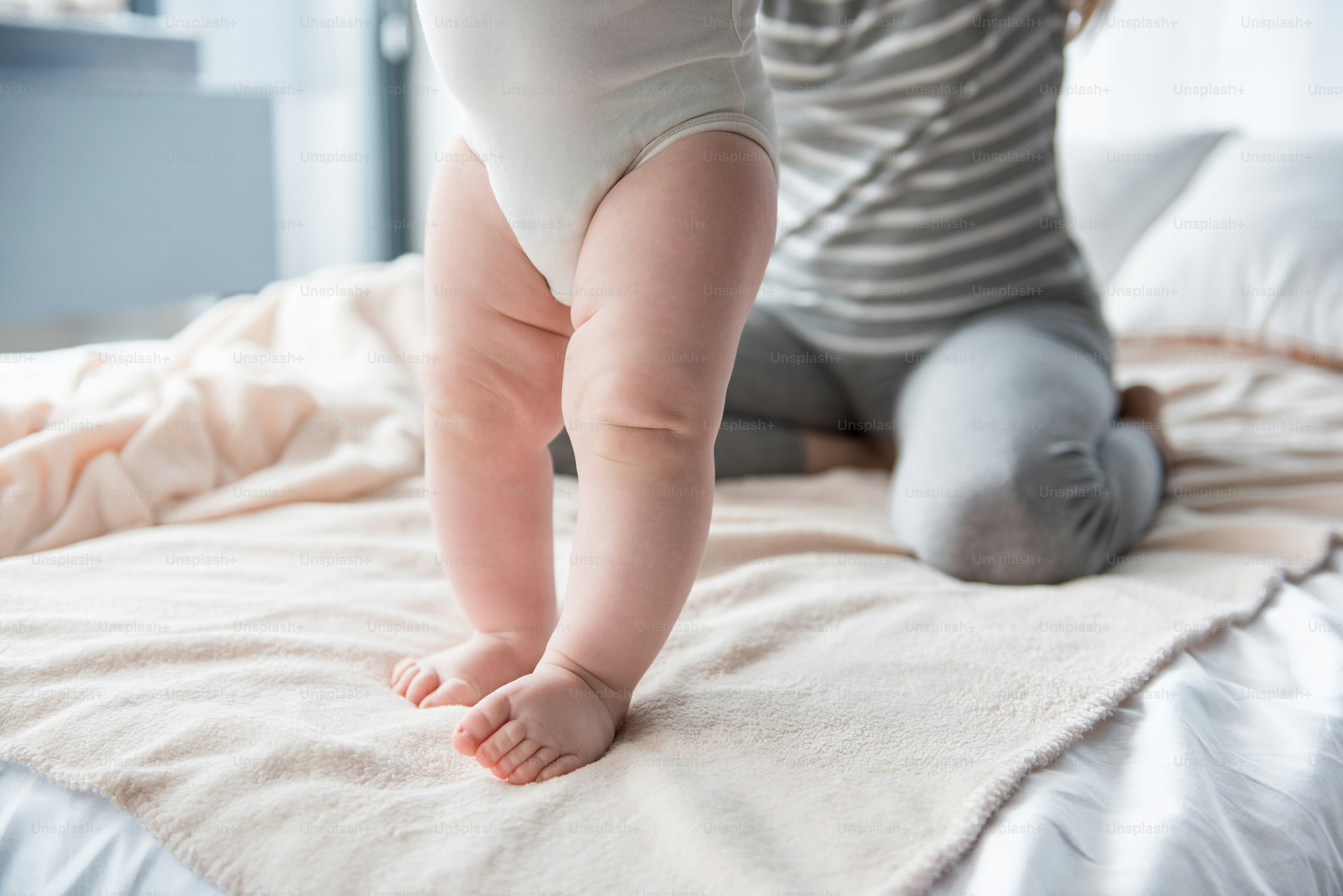 Close up of infant legs standing on bed, female sitting on background ...