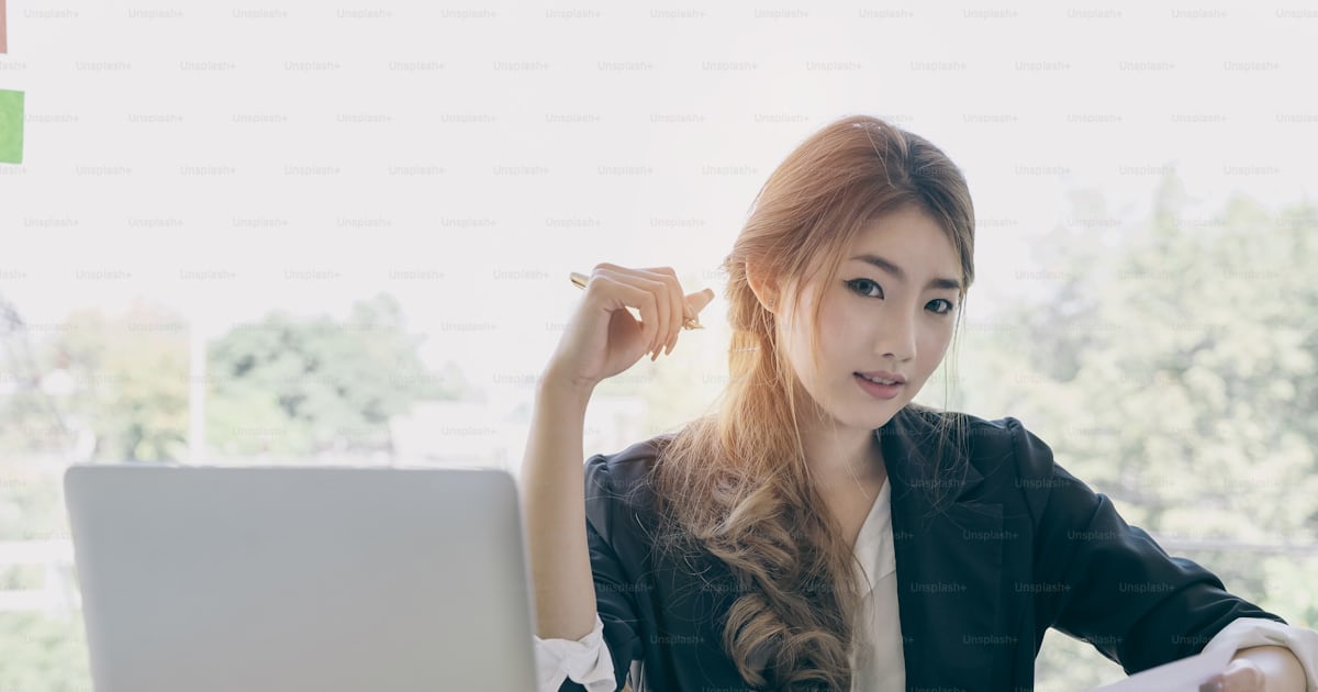 매력적인 아시아 젊은 사업가가 노트북으로 작업하는 동안 밝은 창문이 있는 책상에서 워크스테이션에서 미소를 짓고 있다 사진