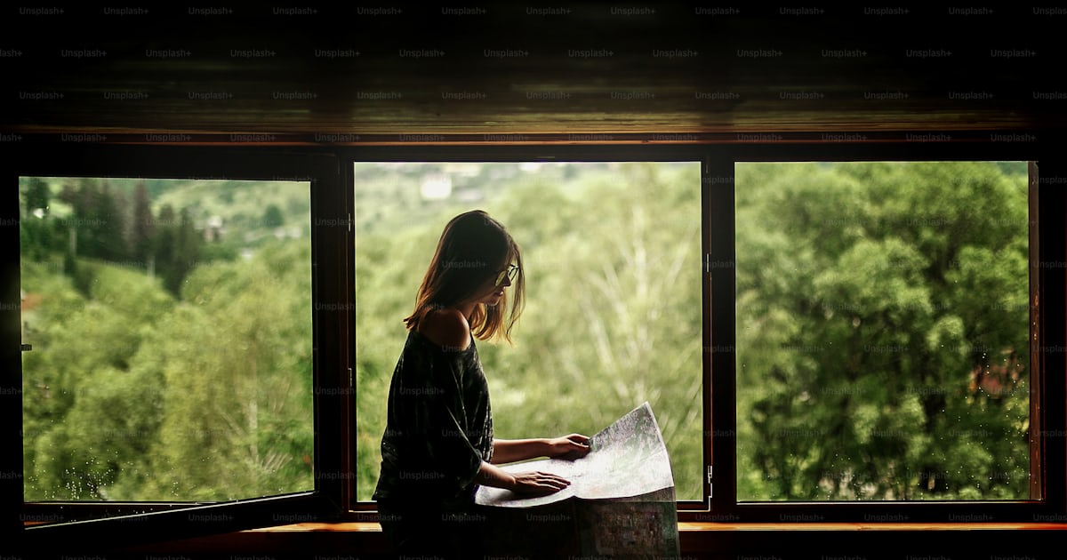 木造家屋の素晴らしい山々を眺めながら窓辺に座って地図を持っている女性、夏の旅行や建築家のコンセプトを計画している女性、テキスト用のスペースの