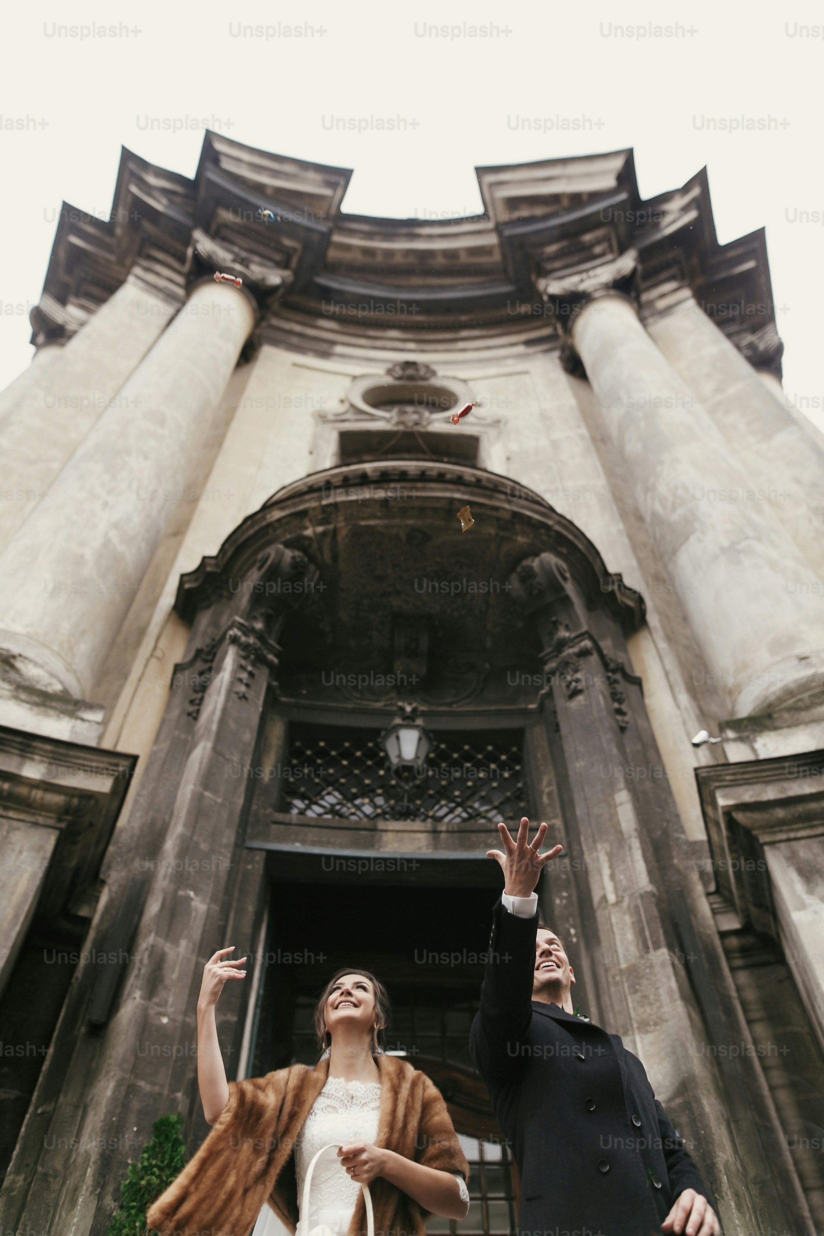 Gorgeous bride in coat and stylish groom throwing candy near church ...