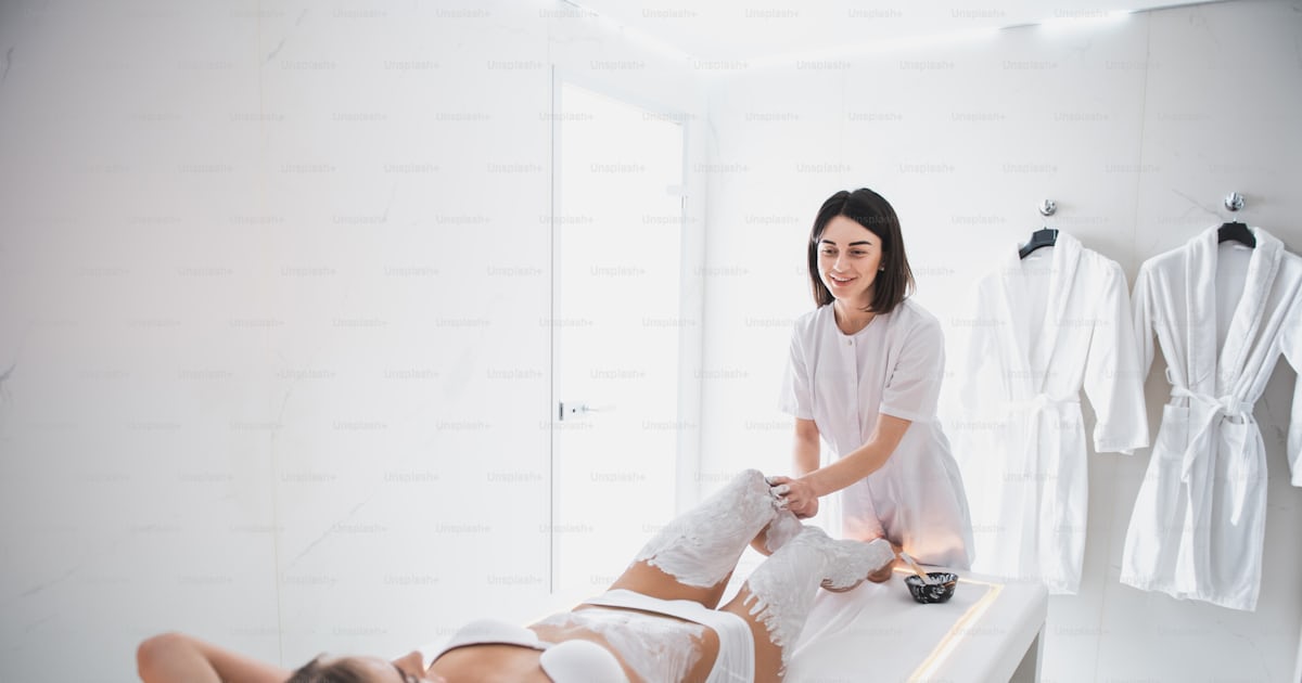 즐거운 절차 흰 속옷을 입은 젊은 아가씨가 마사지 테이블에 누워 스킨케어 절차를 하고 있다 하얀 실험실 코트를 입은 미용사