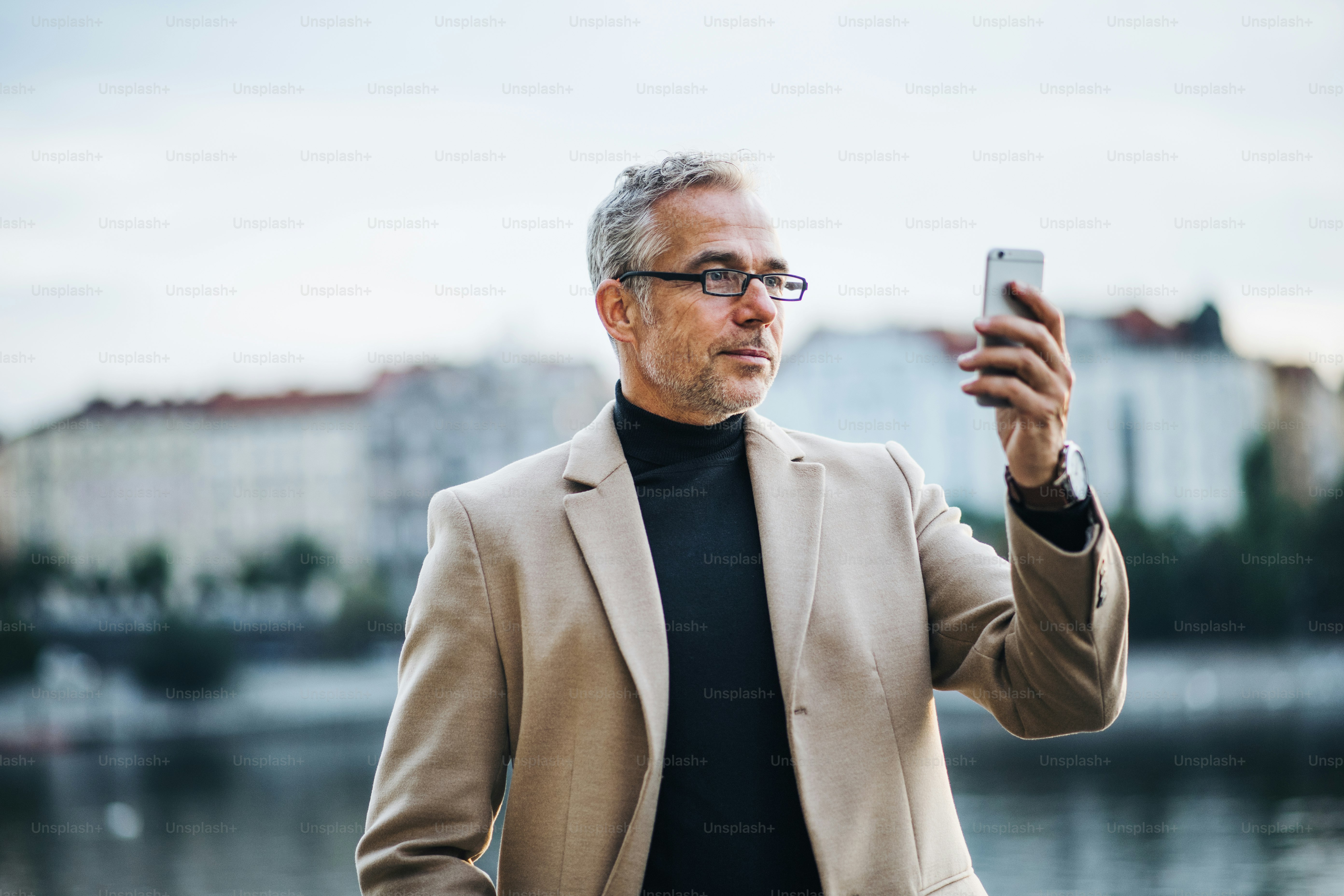 Mature handsome businessman with smartphone standing by river Vltava in ...