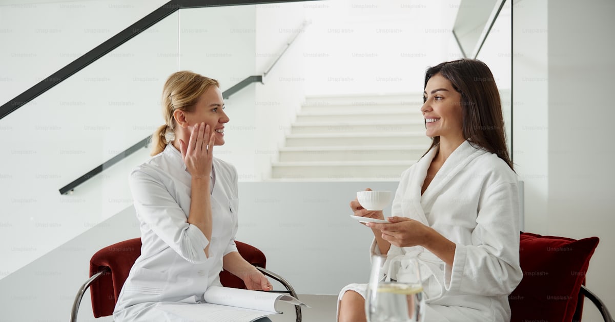 하얀 목욕 가운을 입은 매력적인 여자의 측면 모습은 뜨거운 음료수 한 잔을 들고 미용사와 이야기를 나눴다 그들은 편안한 의자에