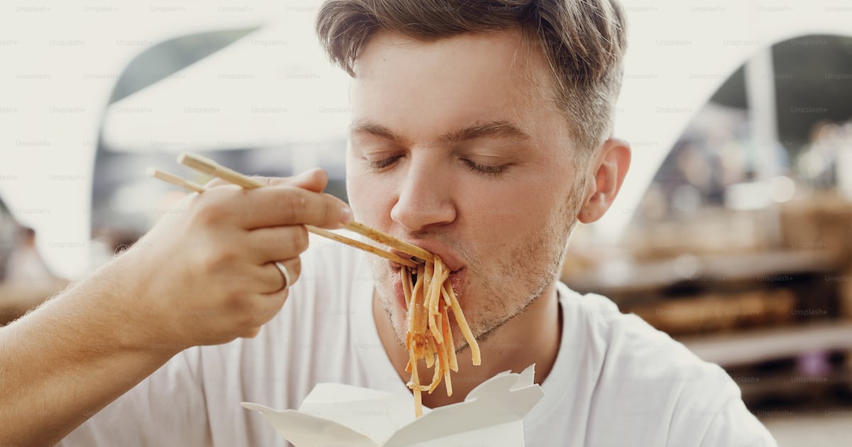 세련된 힙스터 남자는 대나무 젓가락으로 판지 상자에서 야채와 함께 맛있는 냄비 국수를 먹고 있습니다 아시아 거리 음식 축제