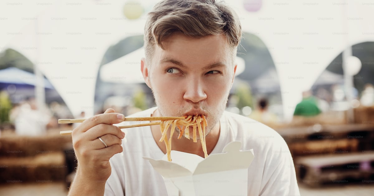 세련된 배고픈 남자는 대나무 젓가락이 달린 판지 상자에서 재미있는 감정으로 맛있는 냄비 국수를 먹습니다 아시아 거리 음식 축제