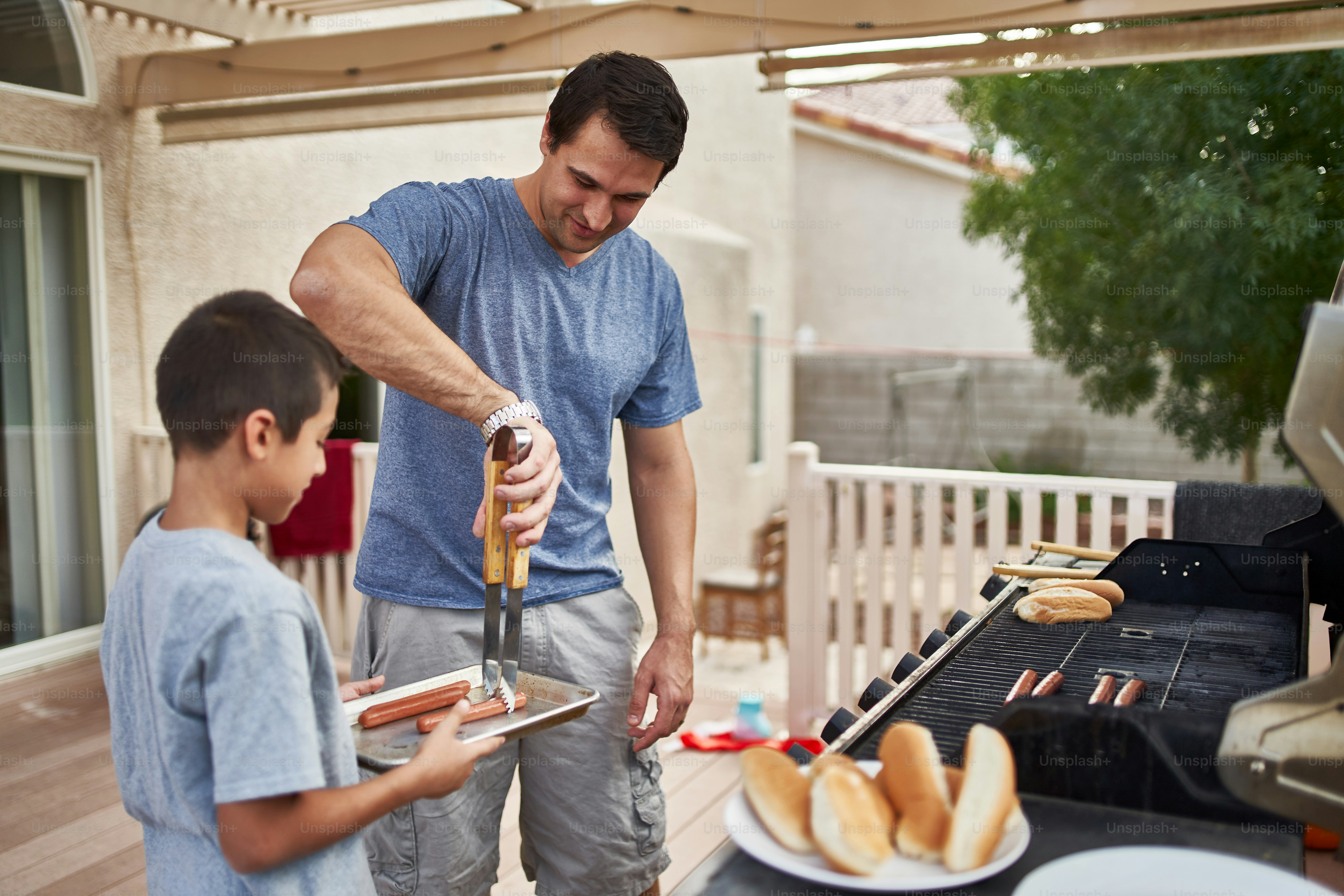 Father and son grilling hot dogs together on backyard gas grill during ...