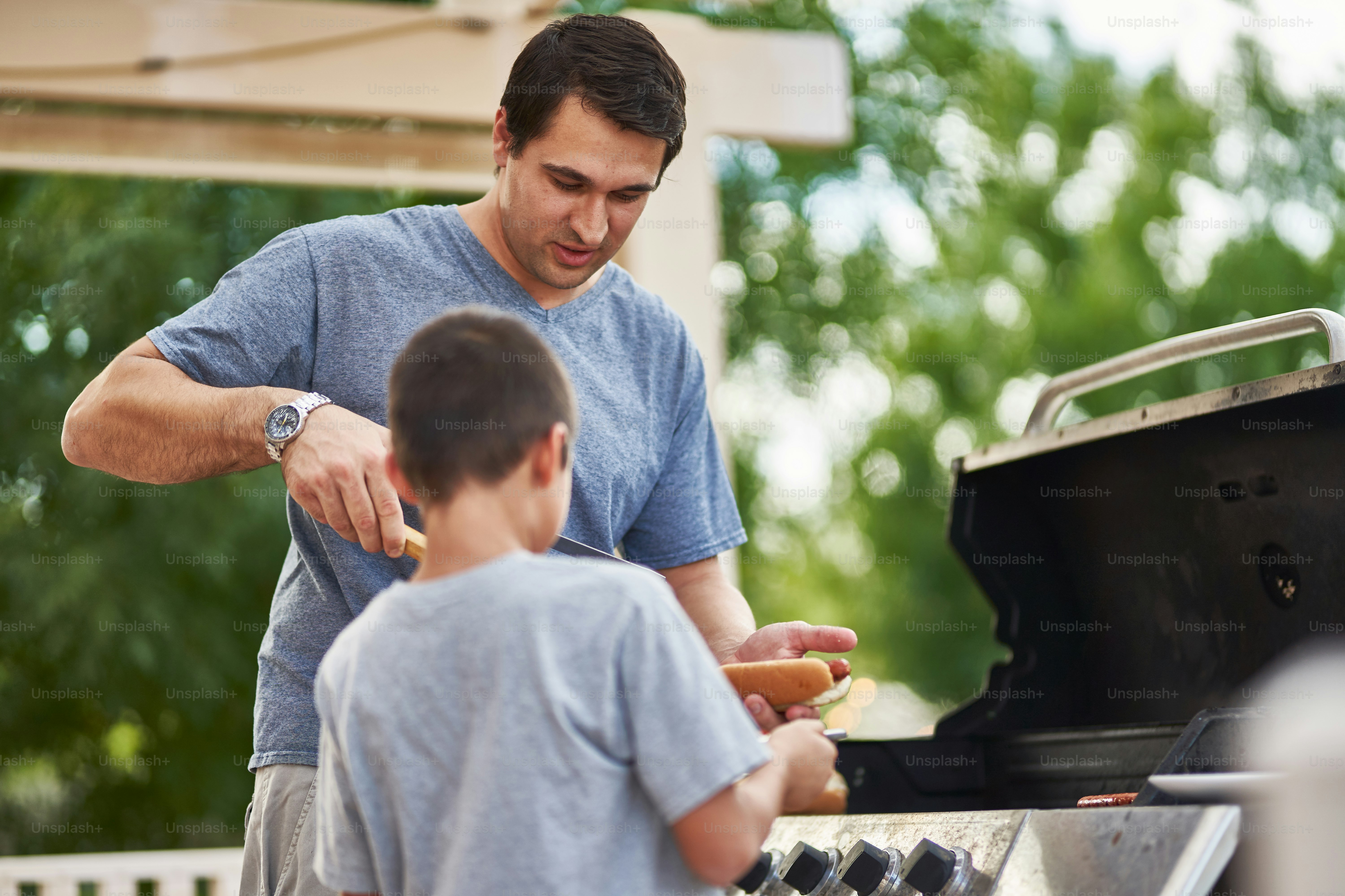 Father and son grilling hot dogs together on backyard gas grill during ...