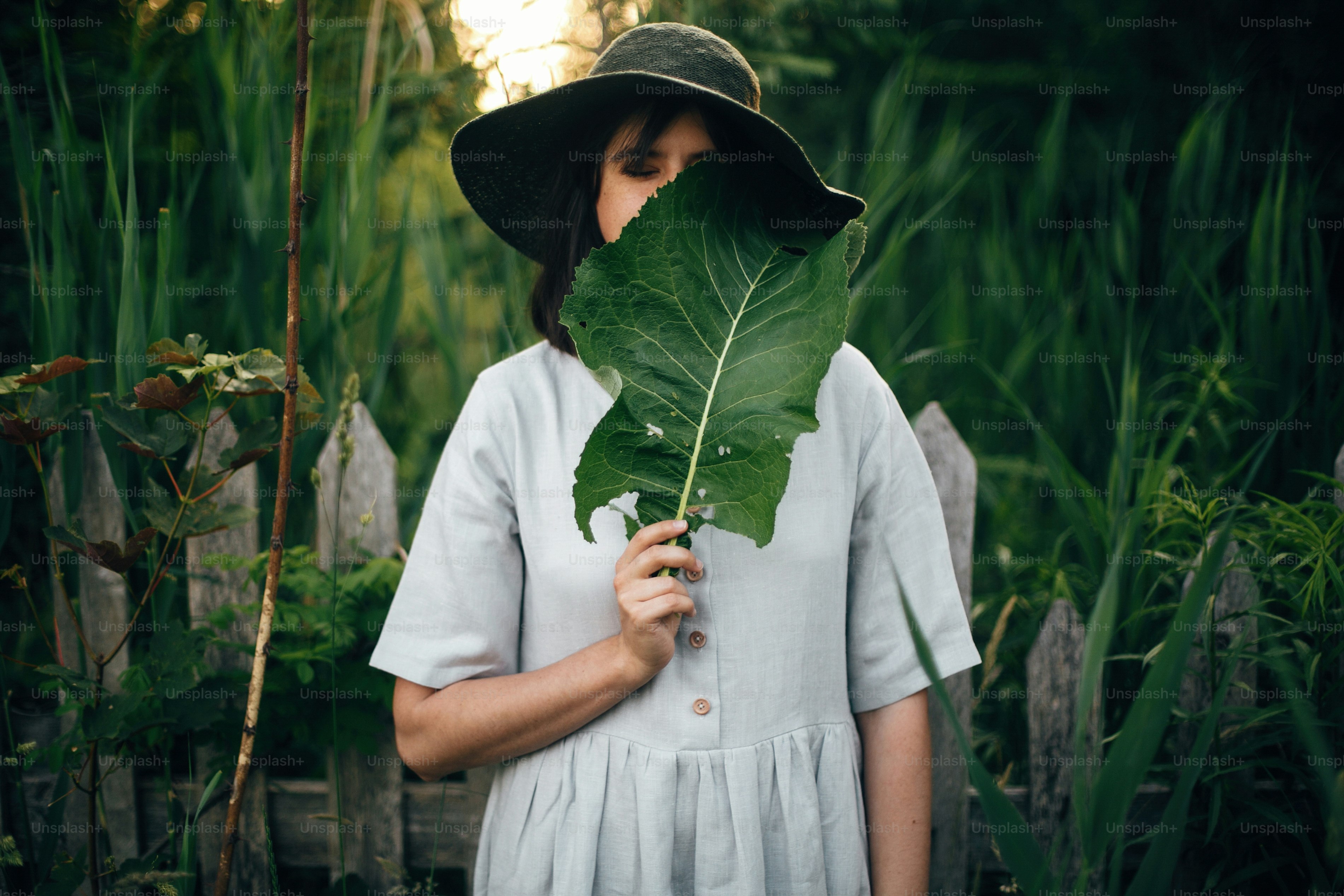 Stylish girl in linen dress holding big green leaf at face at wooden ...