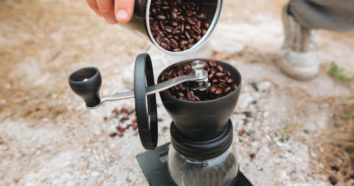 湖の崖でグラインダーに新鮮なコーヒー豆を詰め、キャンプで代替コーヒーを淹れる準備をします。屋外でのピクニックで温かい飲み物を作る。山での