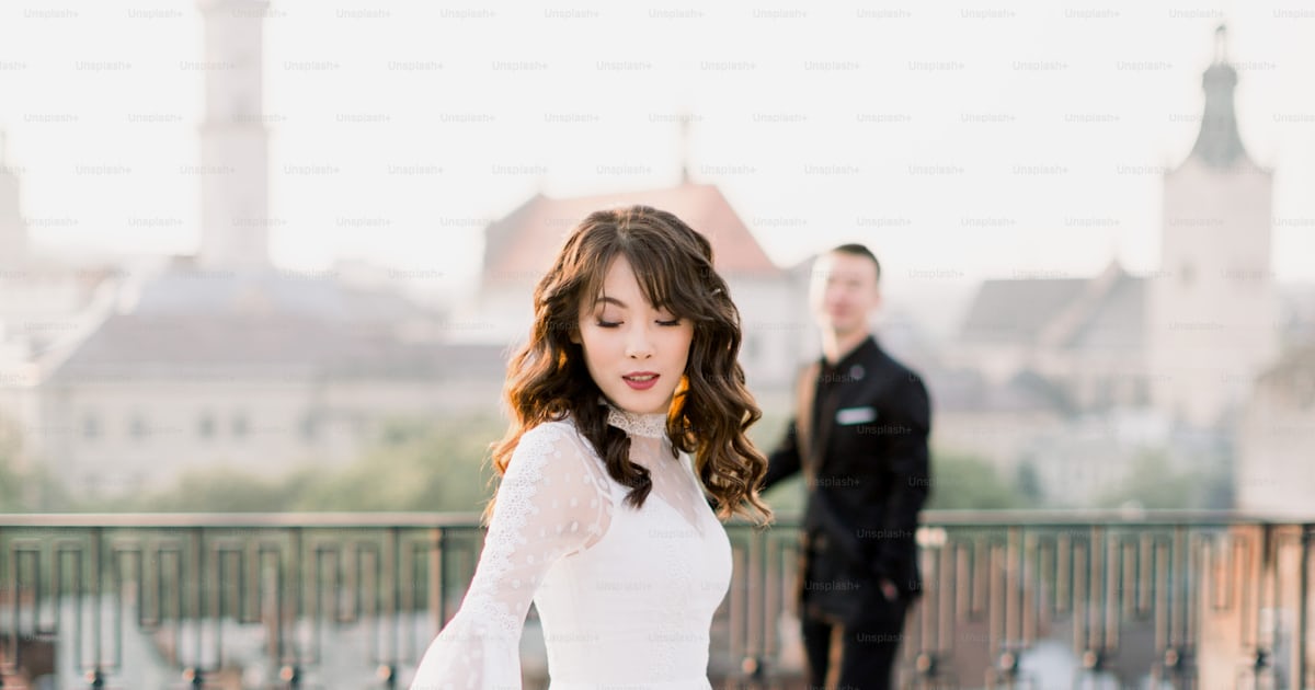 하얀 웨딩 드레스를 입은 젊은 아름다운 아시아 신부가 고대 도시의 테라스에서 춤을 추고 있다 배경에 서 있는 신랑 오래된
