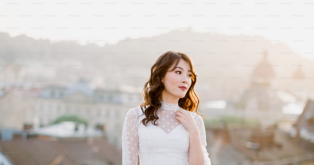 하얀 드레스를 입은 우아한 아시아 여성이 옥상 테라스에 서서 일몰에 옛 유럽 도시의 도시 배경 스카이라인 전망을 보고 있다