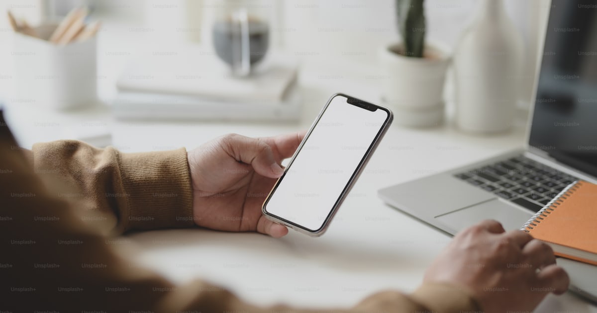 彼の現代の職場で空白の画面のスマート フォンを保持している若い男性フリーランサーのショットをトリミングの写真 タイ Unsplashの写真