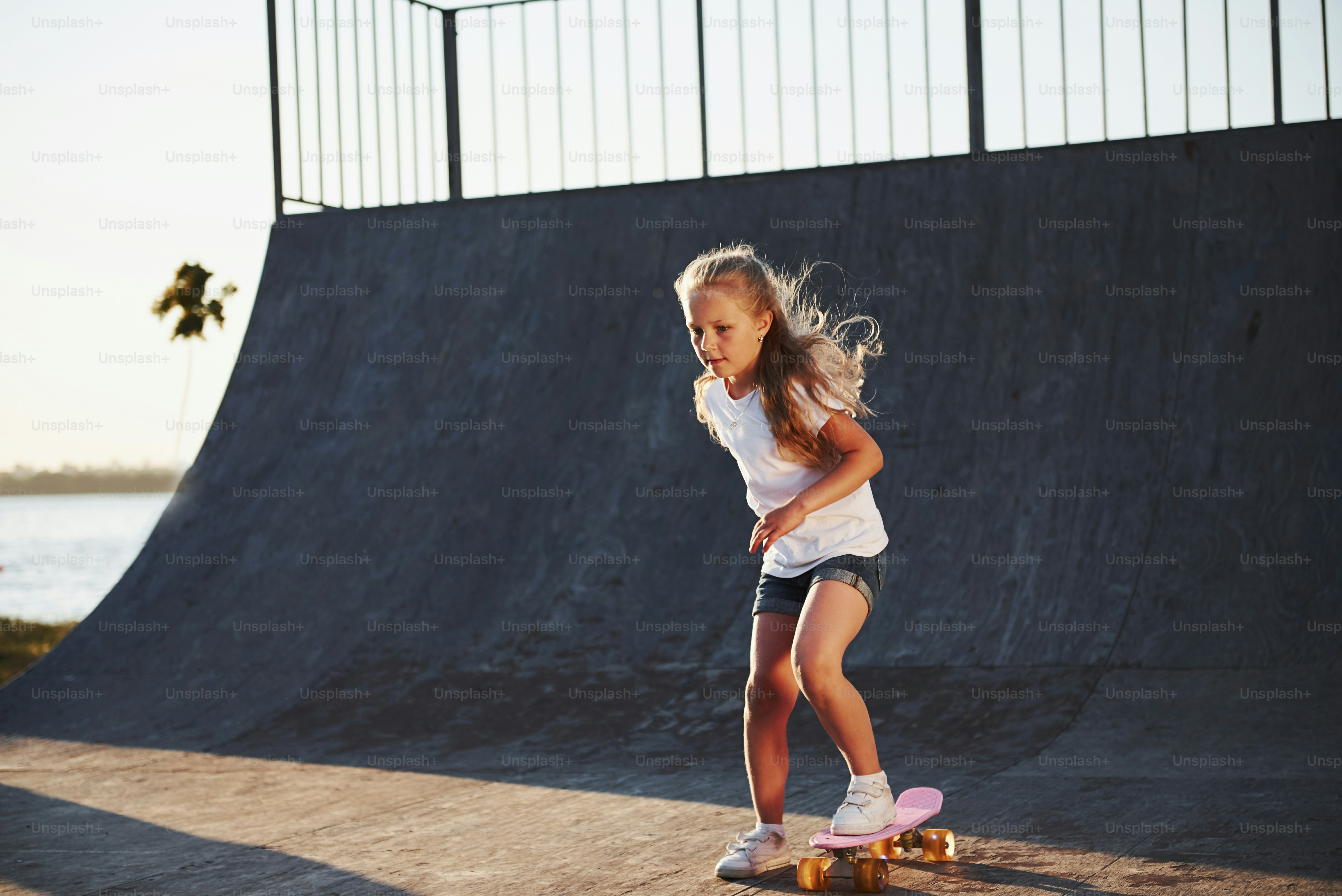 Sunny day. Kid have fun with skate at the ramp. Cheerful little girl ...