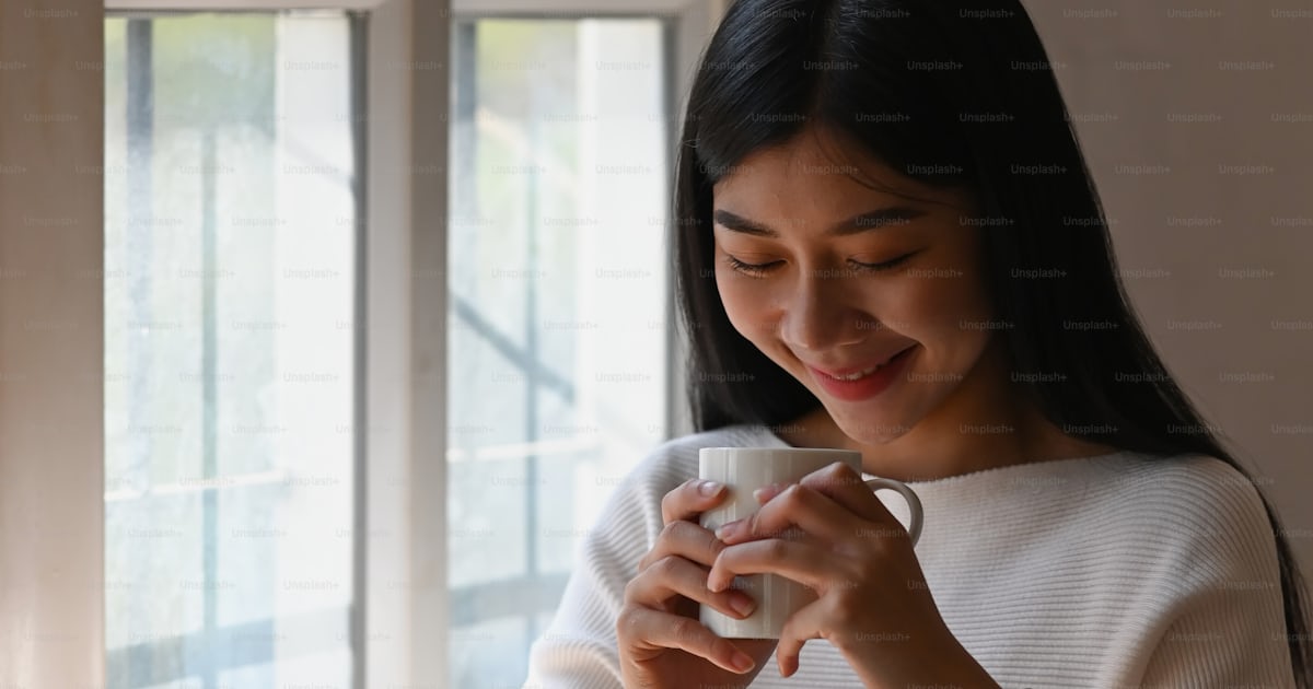 하얀 면 드레스를 입은 젊은 창조적인 여성이 커피 컵을 손에 들고 편안한 거실을 배경으로 나무 테이블에서 일하는 사진 사진