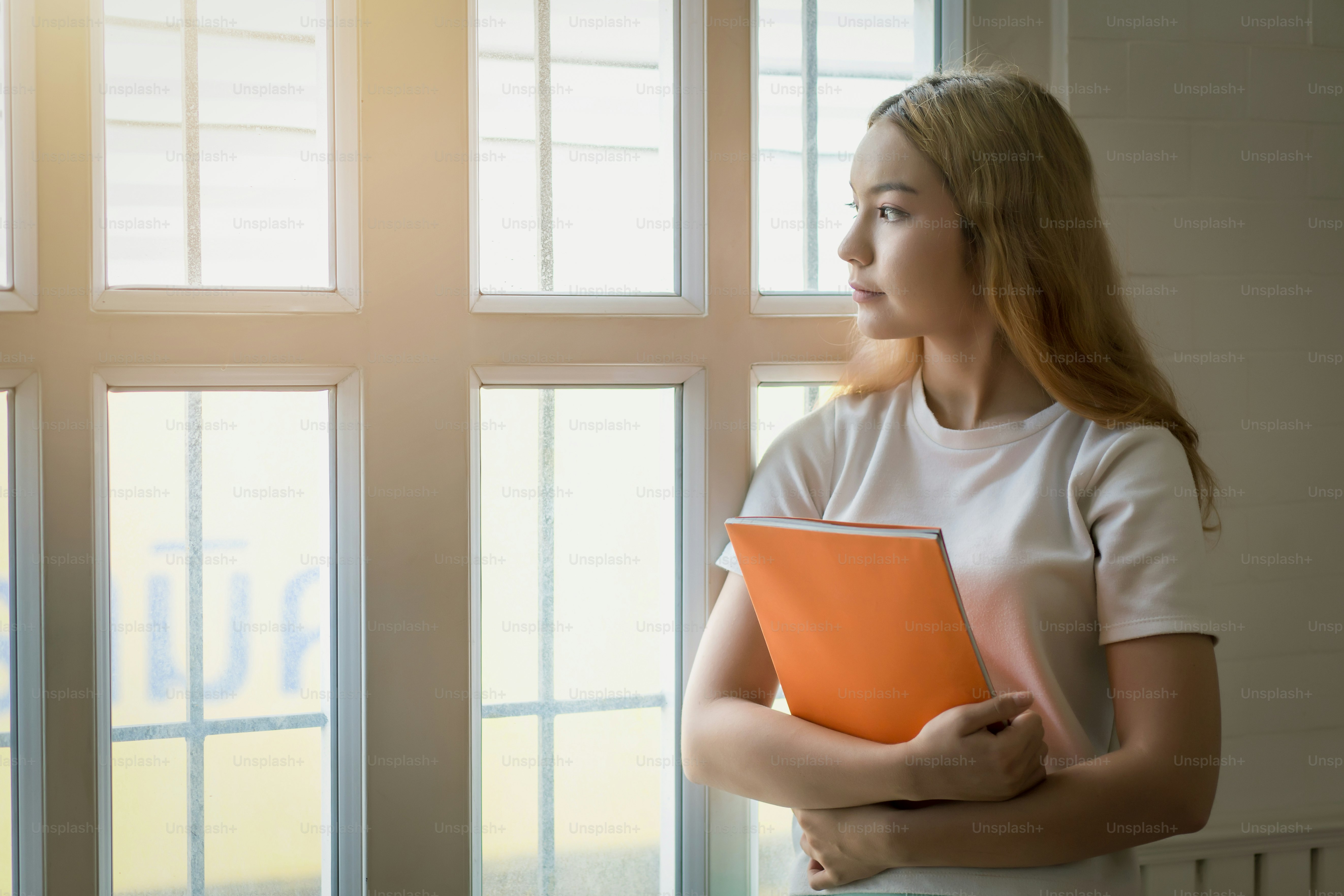 Young asian woman student holding book standing by the window of the ...
