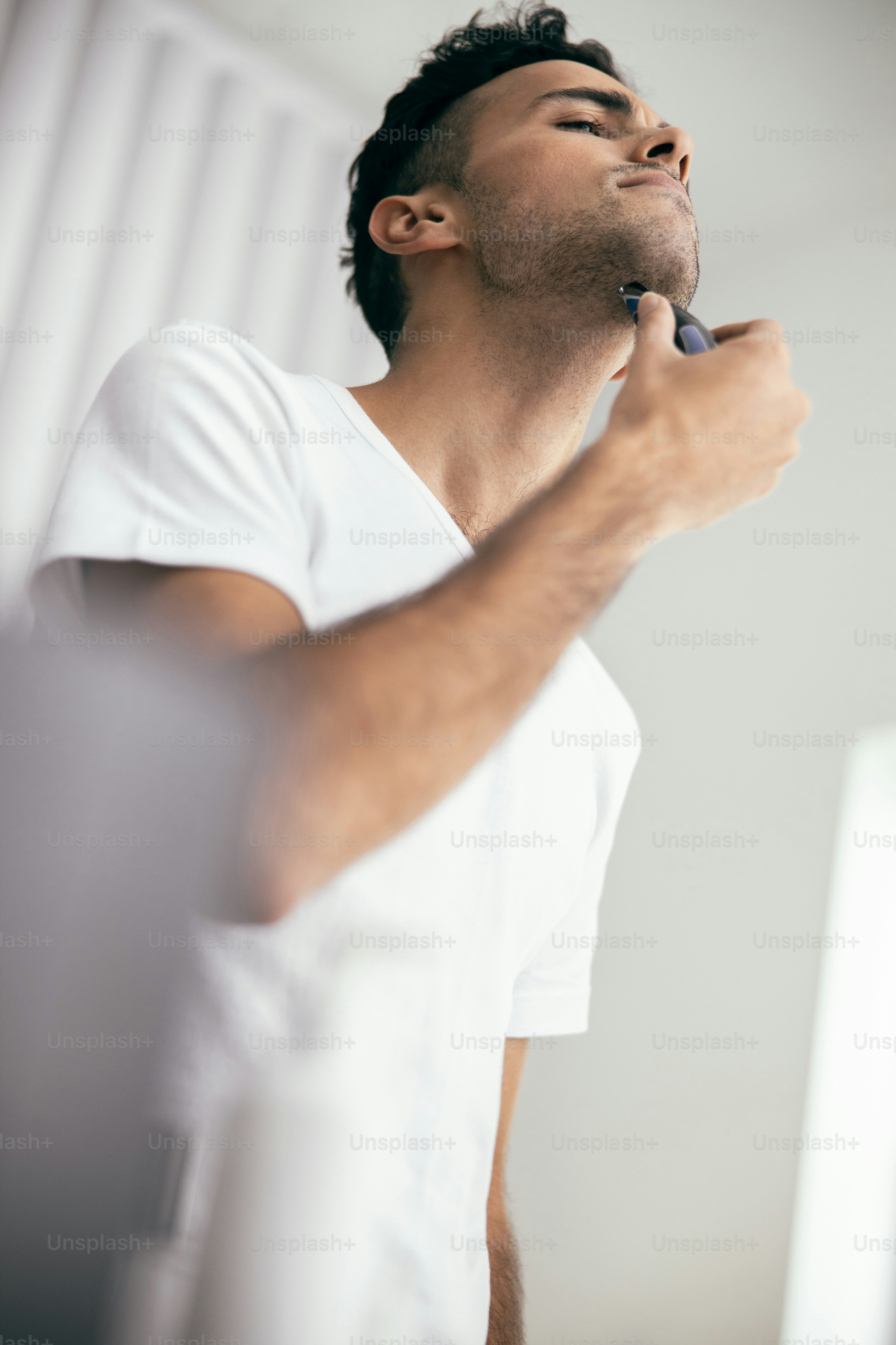 Close up of cheerful guy shaving beard with electric razor at home ...