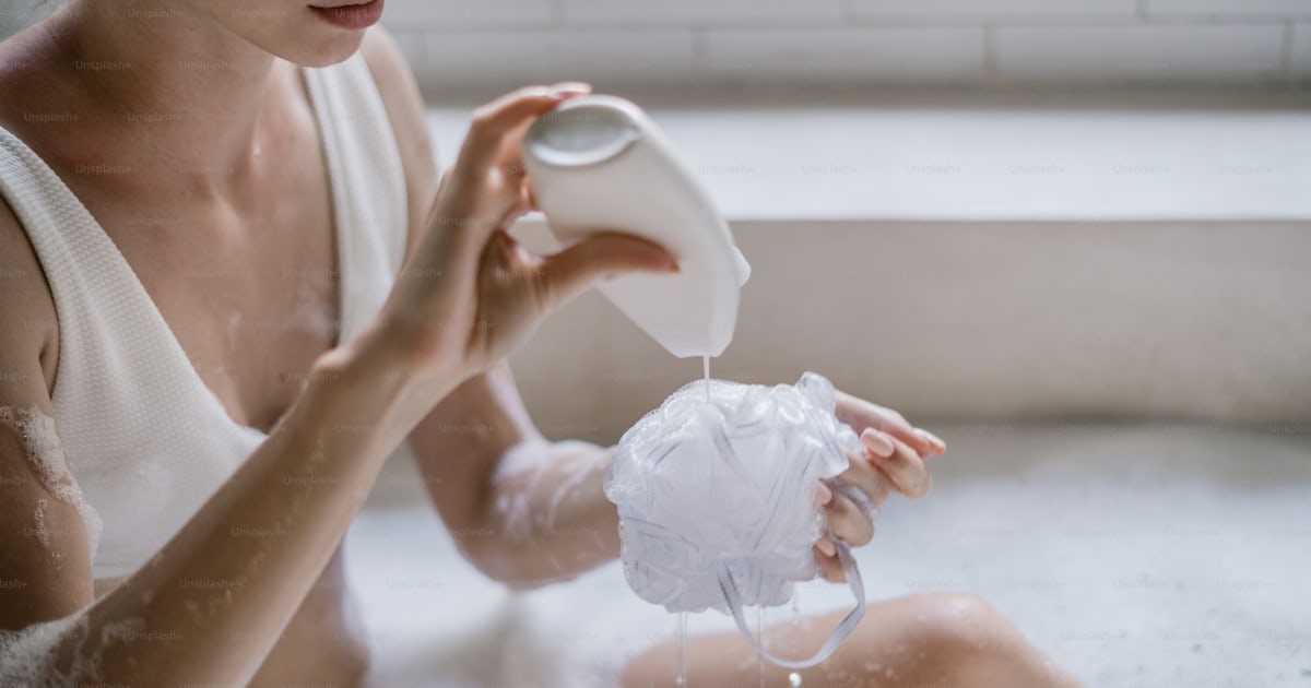 바디 케어 개념입니다 젊은 성인 여성이 스폰지에 샤워 젤을 붓고 목욕을 하고 집에서 비누 거품 물에 누워 있는 모습 사진