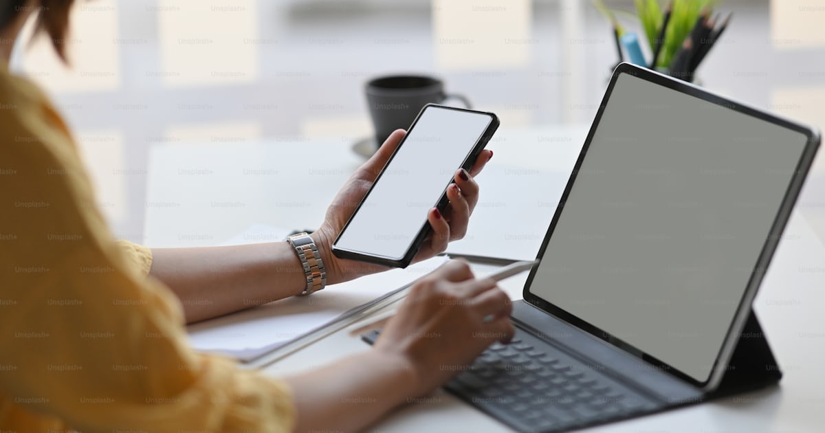 背景として快適なリビング ルームの上の白い作業机に座ってキーボード ケースとコンピューター タブレットを使用しながらスマート フォンと