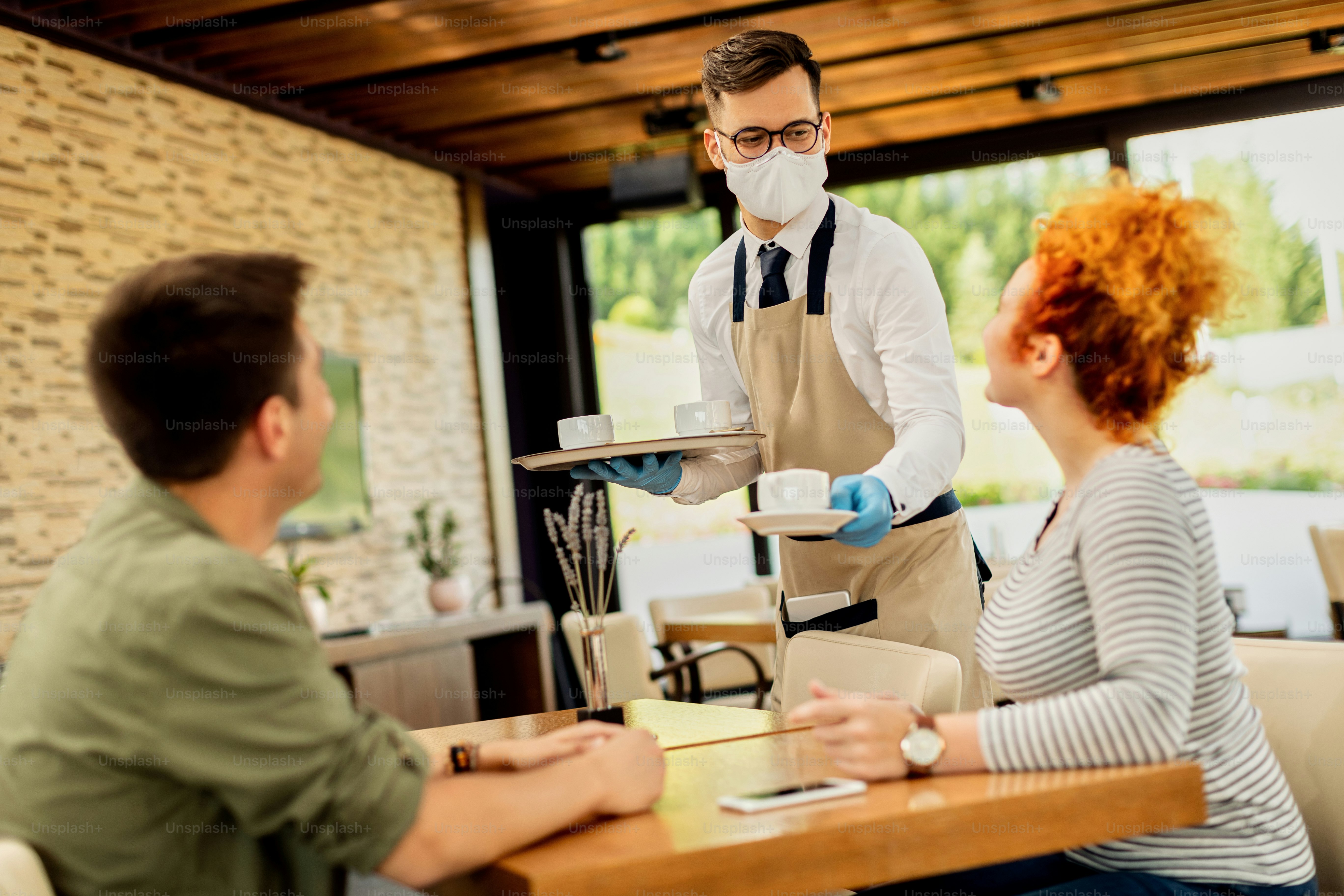 Happy waiter serving coffee to a couple while wearing face mask during ...