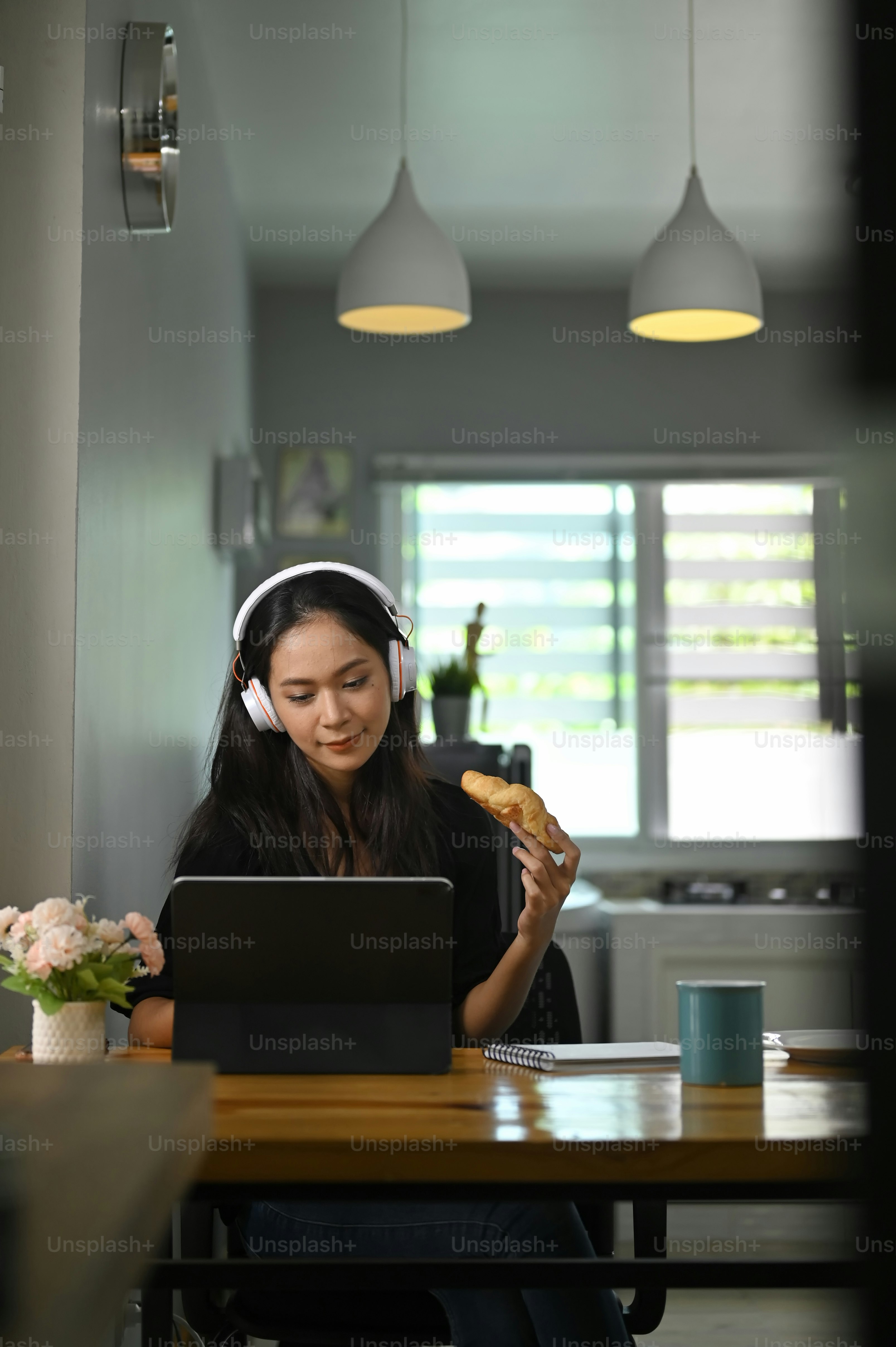 A beautiful woman is eating a croissant while using a computer tablet ...