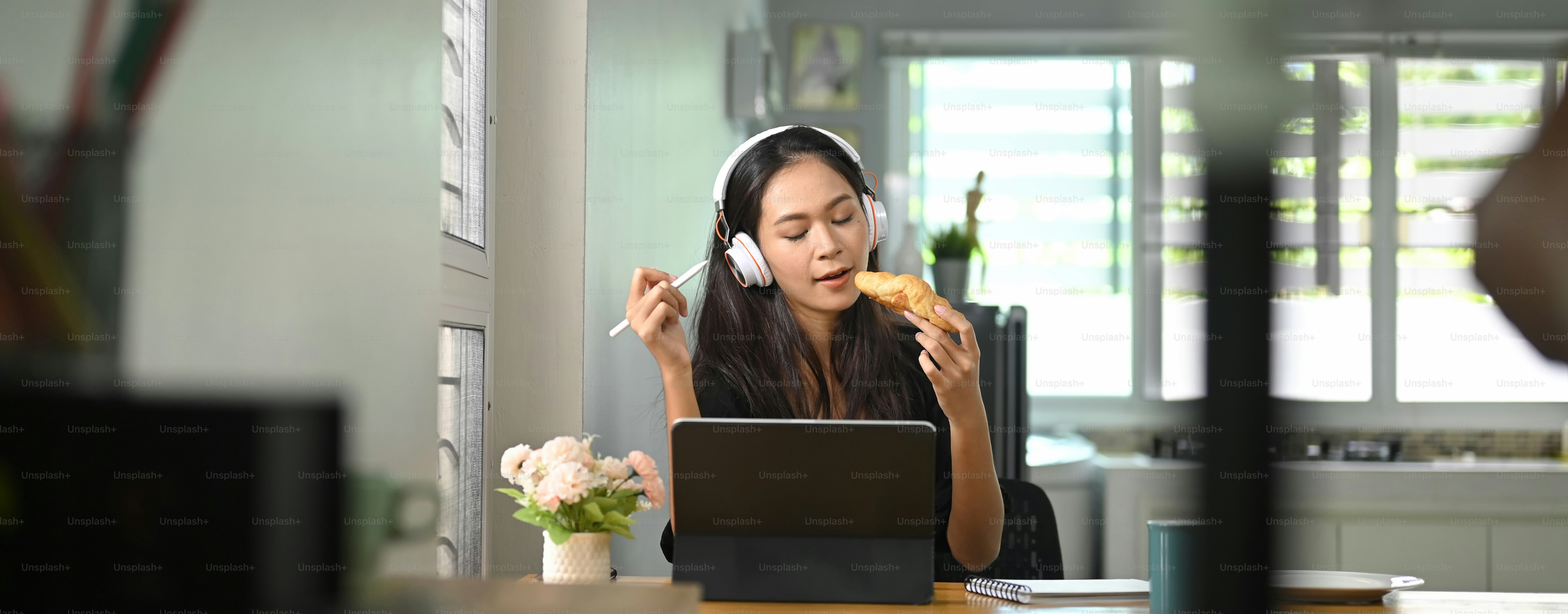 A beautiful woman is eating a croissant while using a computer tablet ...