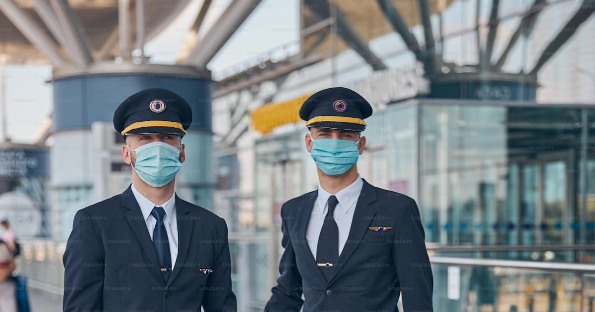 공항에서 비행기를 기다리는 동안 여행 가방을 들고 카메라를 바라보는 보호 안면 마스크를 쓴 잘생긴 젊은 조종사 사진