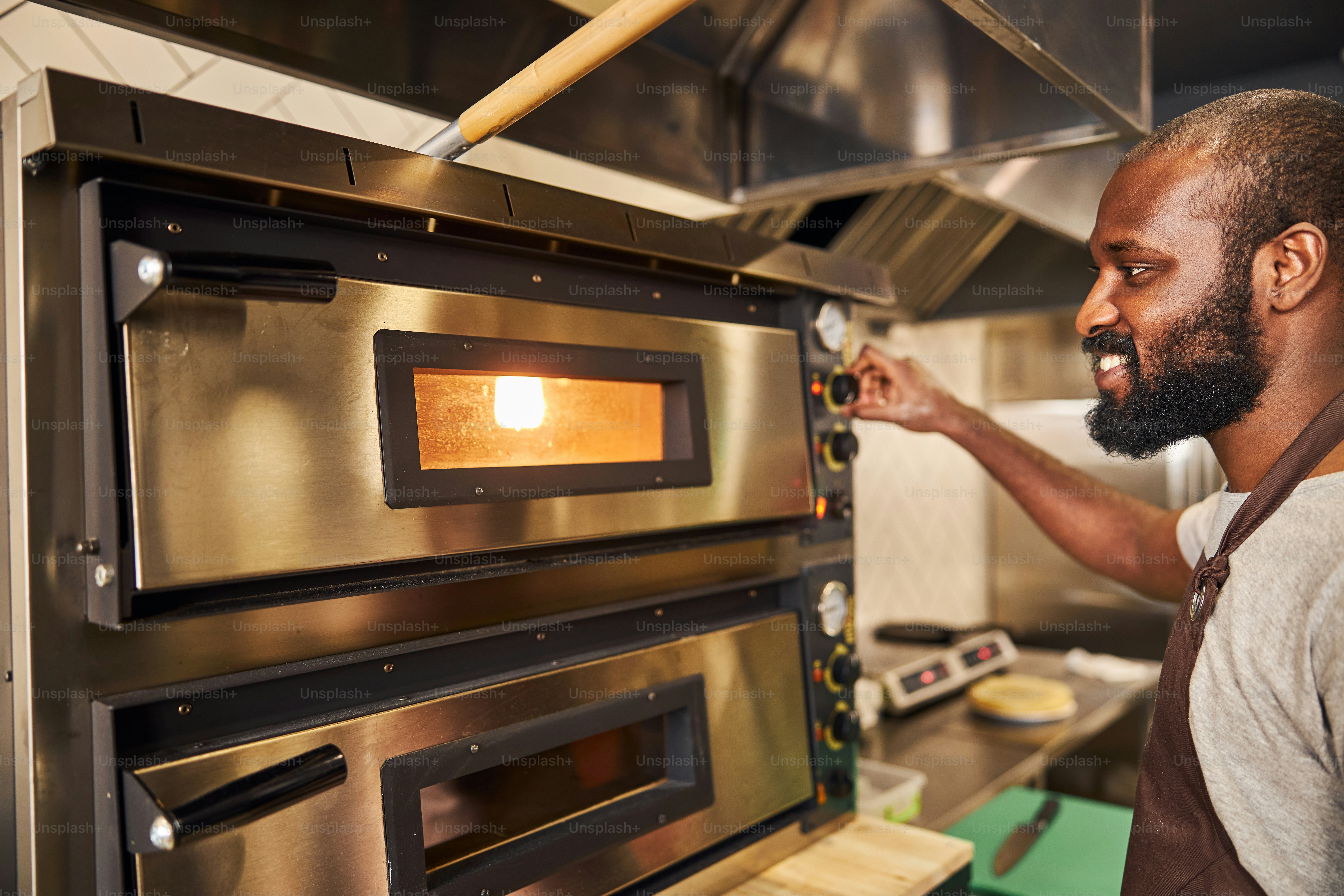 Jolly young handsome guy in apron is standing at oven and looking at ...