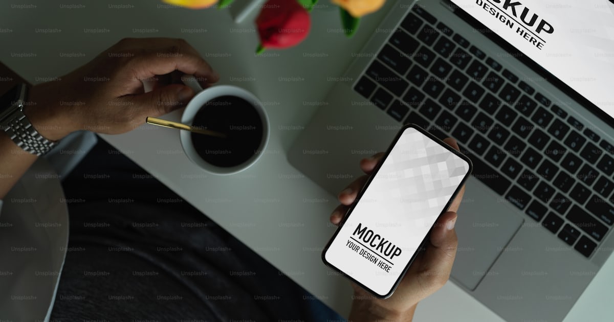作業台にコーヒーカップを持ちながら、空白の画面のスマートフォンのモックアップを保持している男性の上面図、クリッピングパスの写真