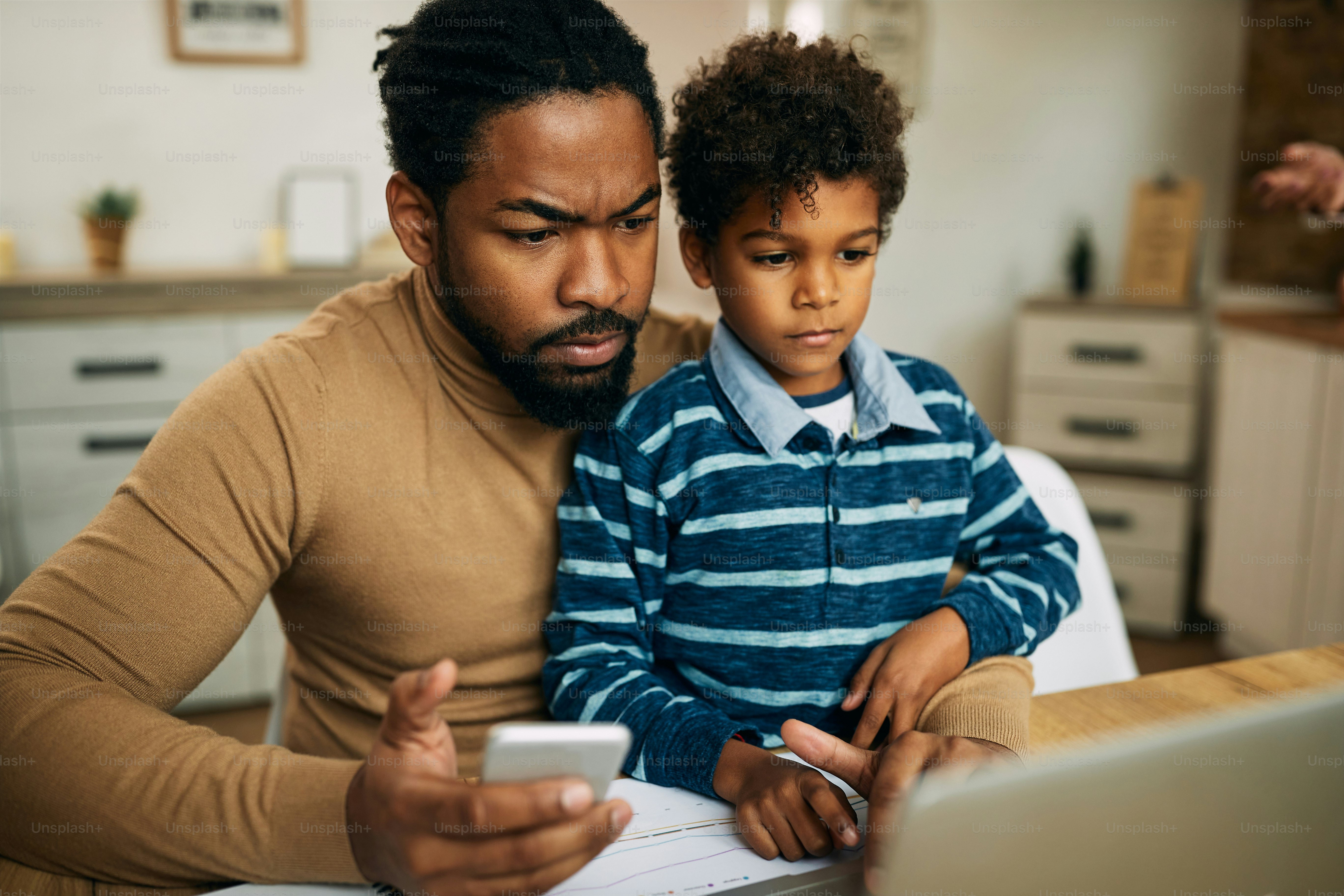 Black working father using computer and smart phone while his son in ...