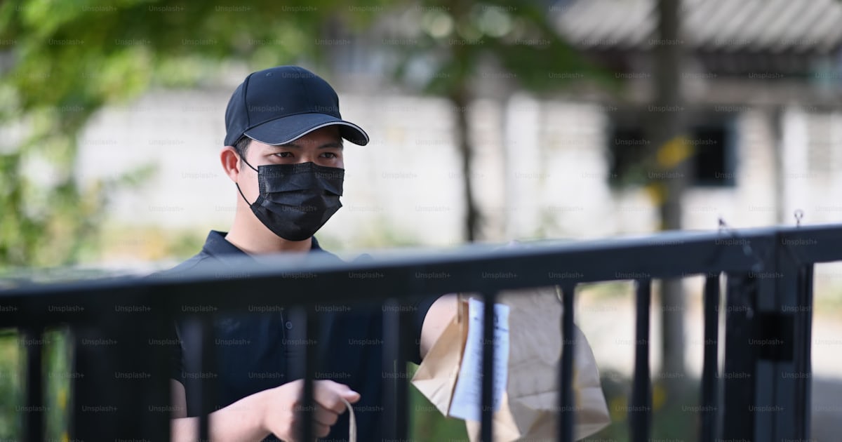 음식이 담긴 종이 봉지를 들고 보호 마스크를 쓴 남자를 배달하고 출입구에서 고객을 기다립니다 음식 배달 서비스 사진