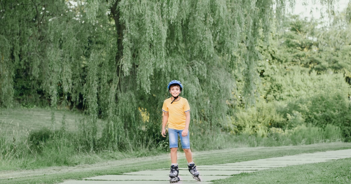 夏の日に公園の道路でローラースケートに乗っているヘルメットの幸せな笑顔の白人の10代の少年。季節の屋外の子供活動スポーツ。健康的な子供時代の