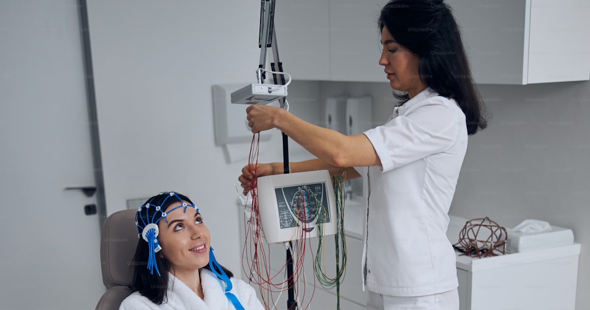 테리 목욕 가운을 입고 자격을 갖춘 신경 생리학자가 뇌파 검사를 위해 뇌파계를 설정하는 것을 지켜보는 웃는 환자 사진
