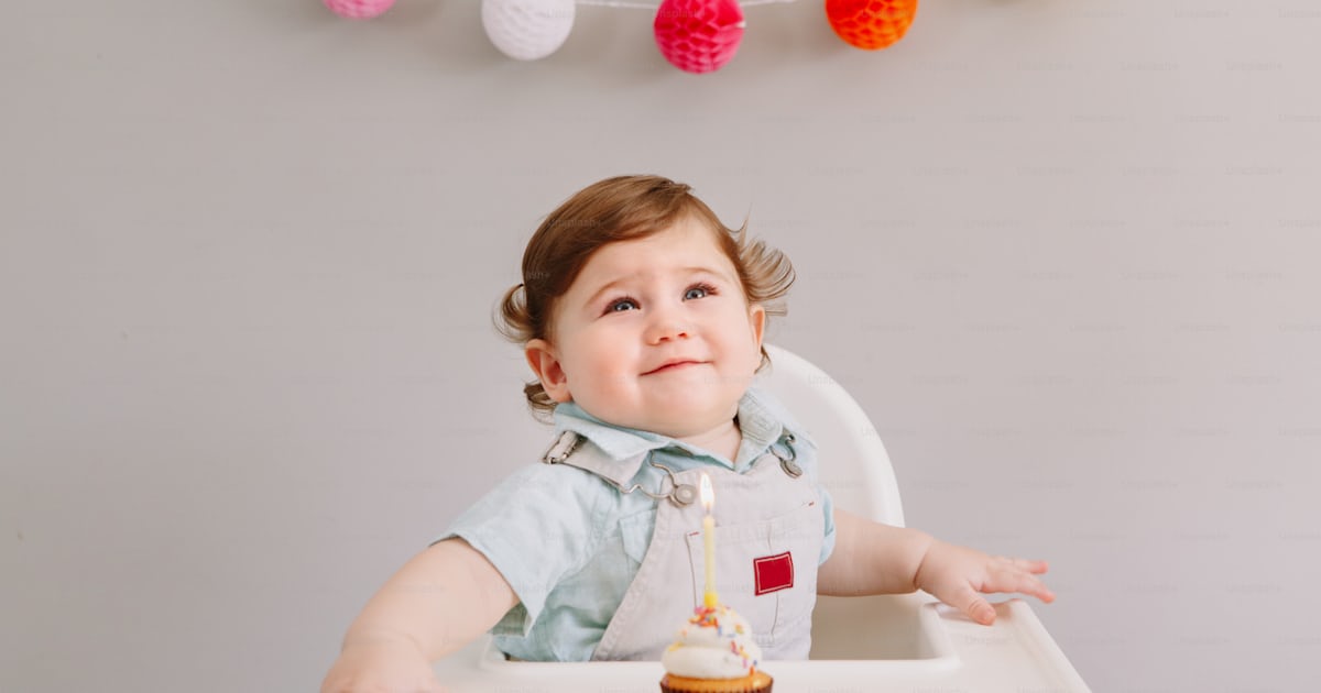 집에서 첫 생일을 축하하는 귀여운 백인 아기가 웃고 있다 높은 의자에 앉아 촛불이 켜진 맛있는 컵케이크 디저트를 먹는 아이