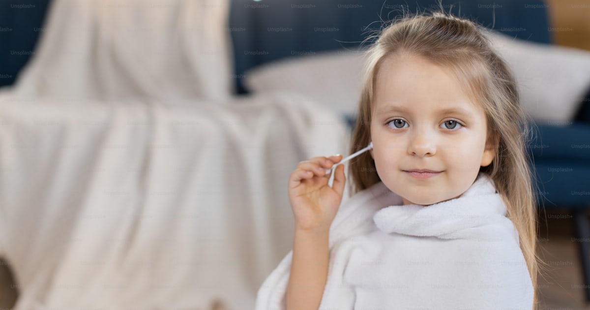 幸せな笑顔の3歳の子供の女の子の屋内ポートレートをクローズアップし、笑顔でカメラに向かって自宅でポーズをとり、綿棒で彼女の小さな耳を掃除します