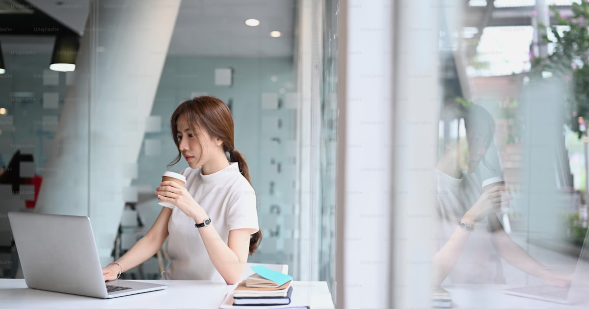 차분한 비즈니스 여성이 창문 근처의 현대적인 직장에 앉아 커피 컵을 들고 노트북 컴퓨터로 작업하고 있다 사진