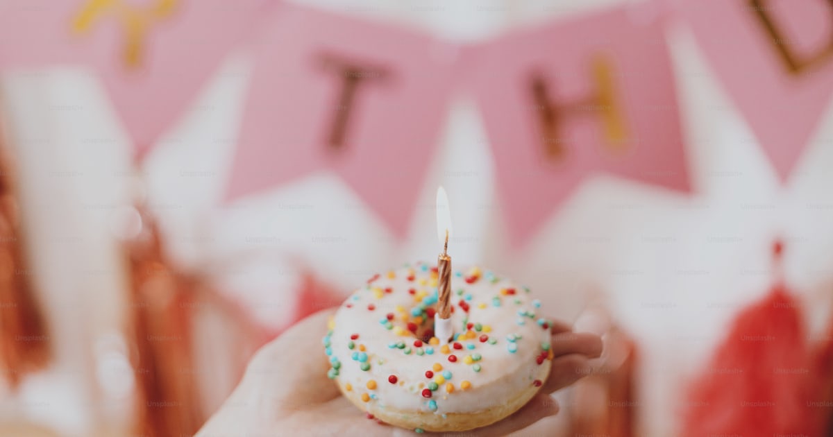 생일 축하해요 분홍색 화환과 방의 장식을 배경으로 촛불이 있는 맛있는 생일 도넛을 들고 있습니다 생일 파티 축하 스프링클이