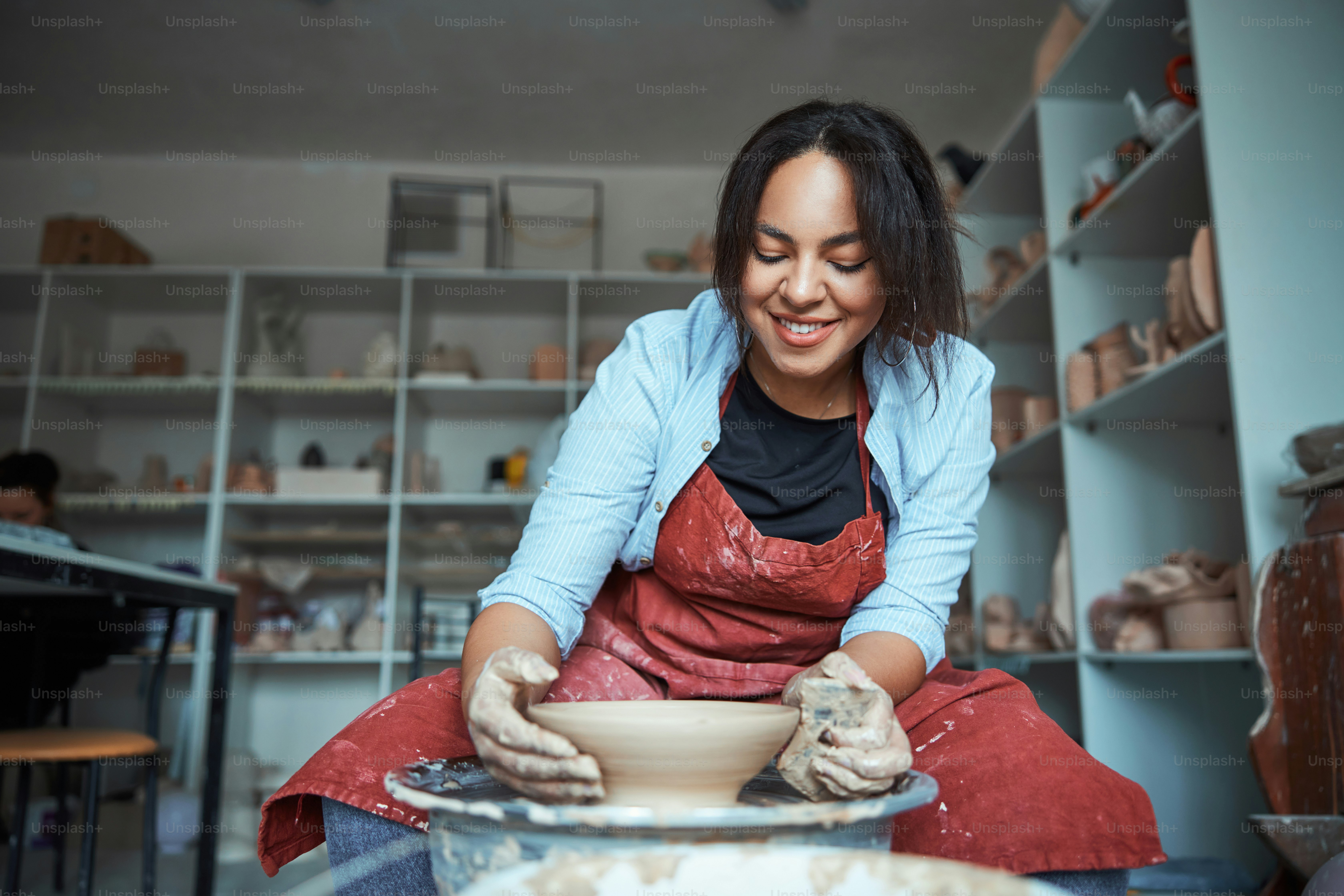 Smiling female potter in apron using grinding block while making clay ...