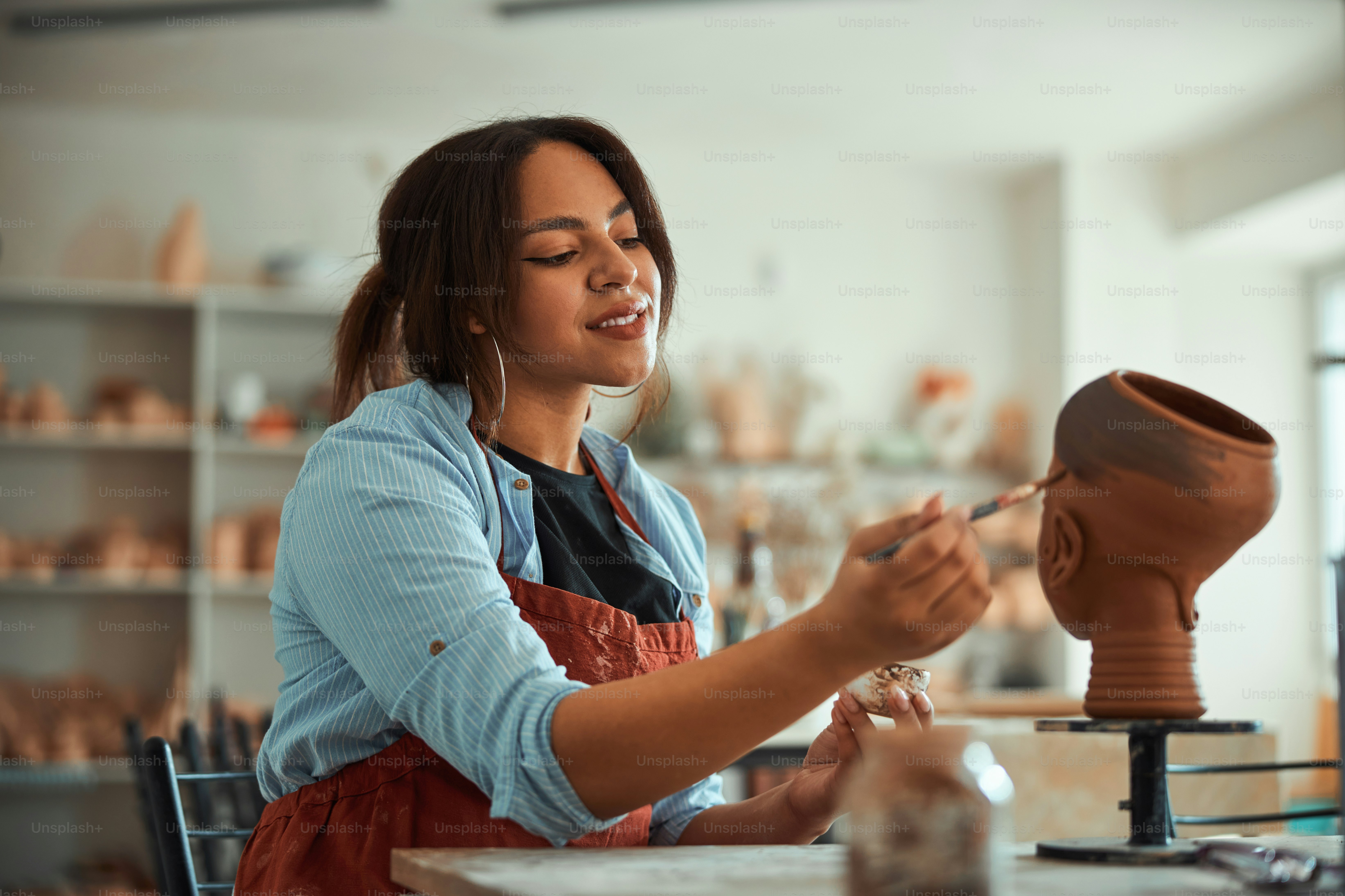 Charming female ceramist painting pottery with paint brush and smiling ...
