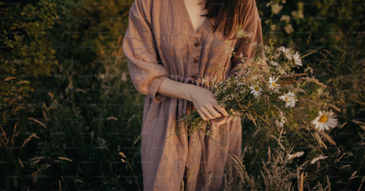 晴れた夕方の夏の牧草地で野生の花を集めているリネンのドレスを着た美しい女性、接写。雰囲気のある瞬間。田舎で花を摘む素朴なドレスを着た