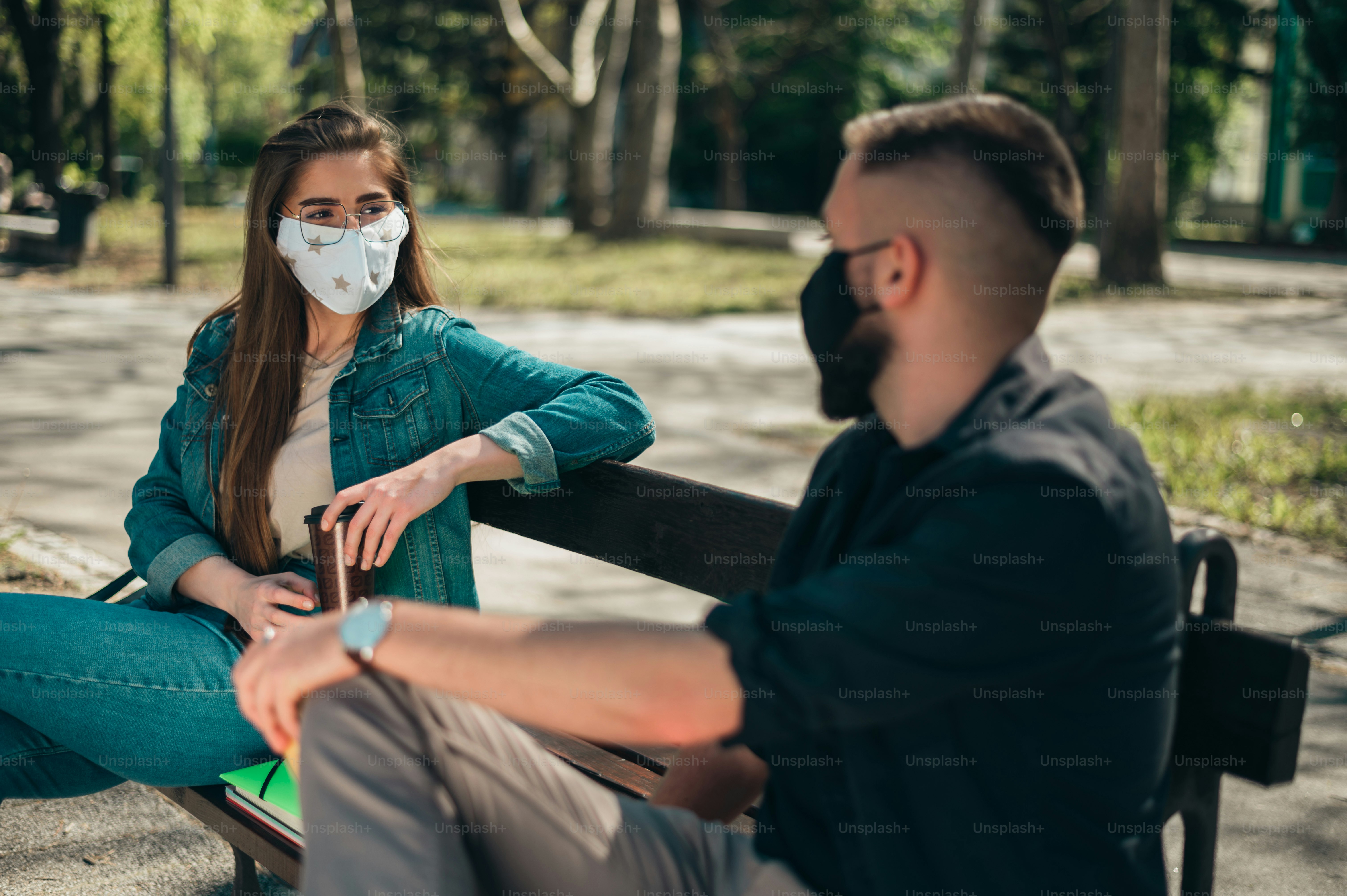 Two young students wearing protective masks and sitting on a bench in a ...