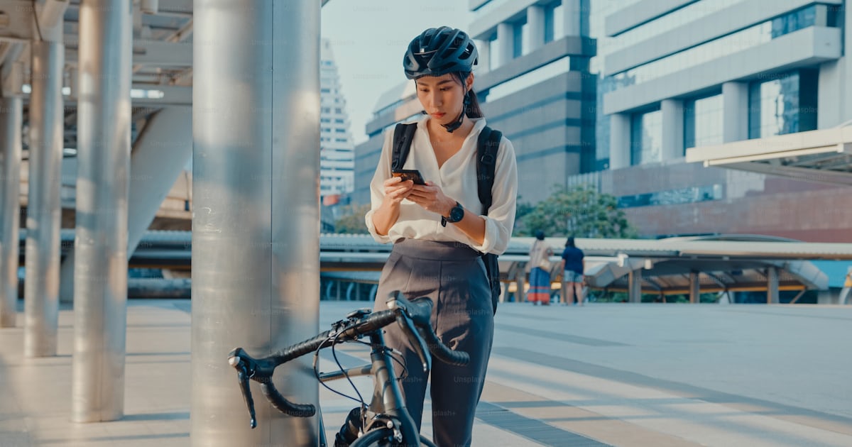 バックパックを背負った笑顔のアジアの実業家は、自転車で通りの街のスタンドでスマートフォンのルックカメラを使用してオフィスに出勤します。スポーツ