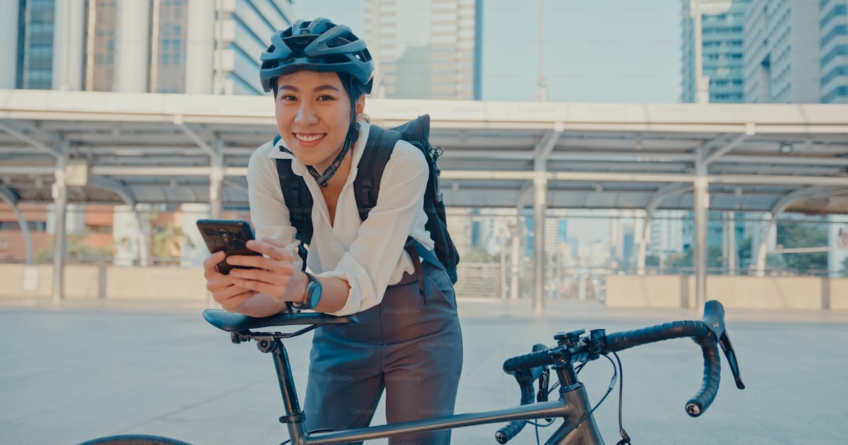 バックパックを持った笑顔のアジアのビジネスウーマンは、オフィスで仕事に行く自転車で街のスタンドでスマートフォンの外観のカメラを使用します