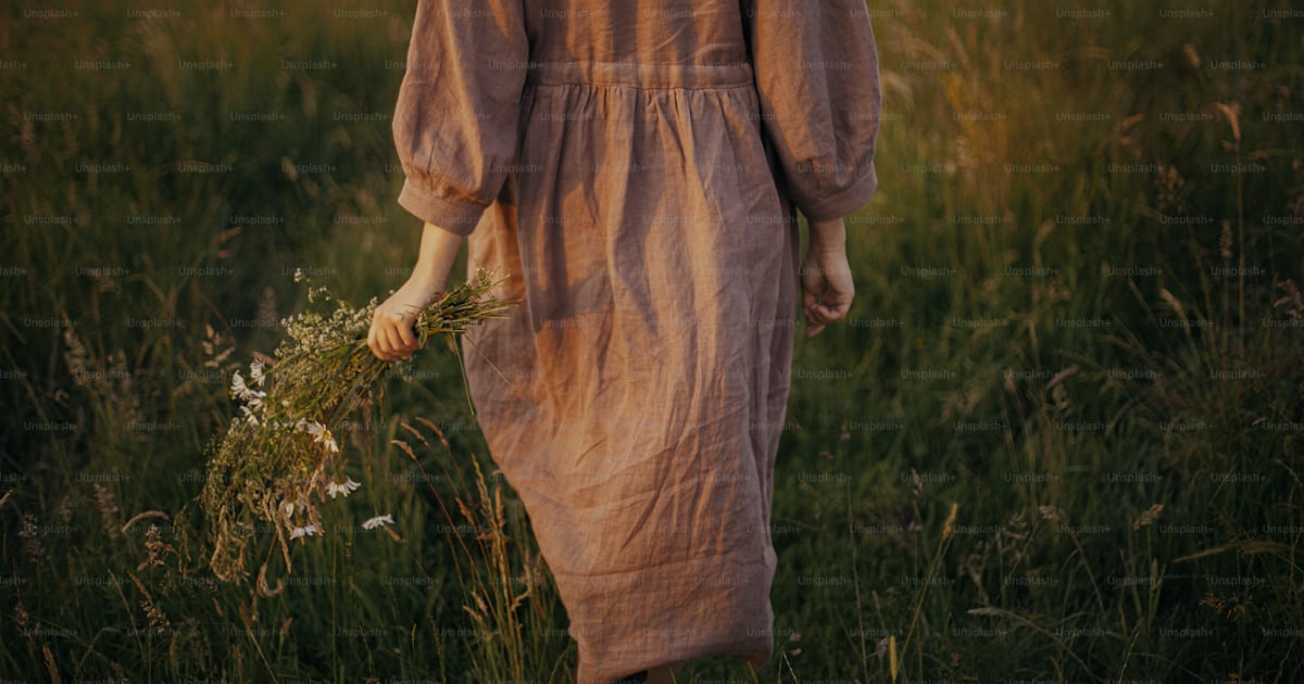 夕暮れの夏の牧草地で野生の花を手に歩いているリネンのドレスを着た美しい女性、後ろ姿。田舎で夜にリラックスした素朴なドレスを着た若い女性。大気の