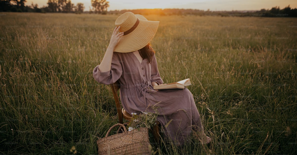 夕暮れの夏の牧草地で素朴な椅子に座って本と花のバスケットを持つ帽子とリネンのドレスを着た美しい女性。雰囲気の穏やかな瞬間。田舎で本を読む若い