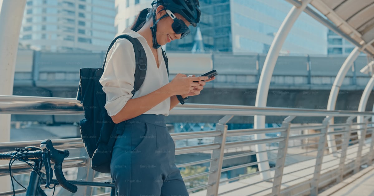 バックパックを持った幸せなアジアのビジネスウーマンは、自転車で通りの街のスタンドでスマートフォンを使用し、オフィスで仕事に行きます。スポーツの