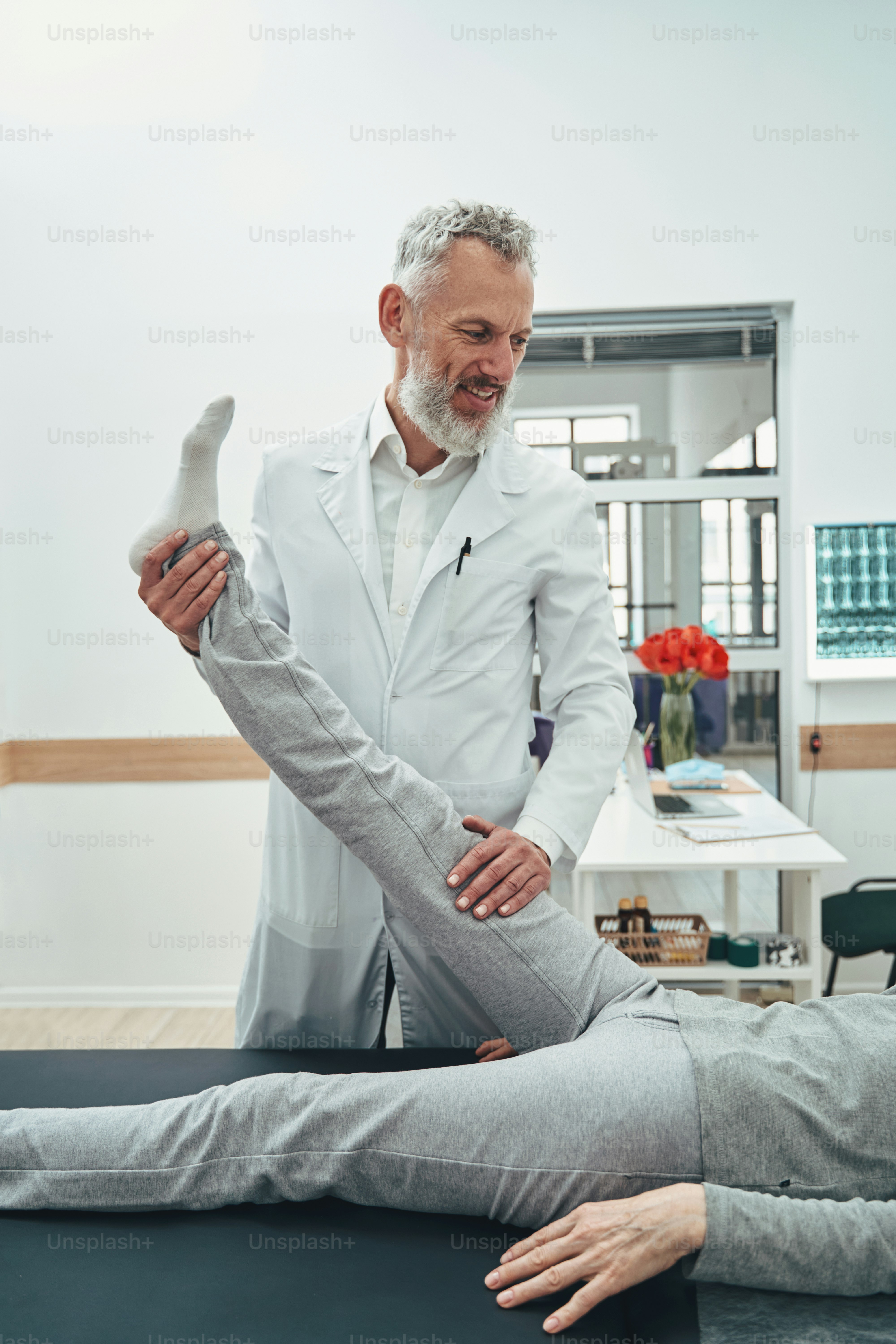 Male doctor lifting an extended lower limb of a patient lying in a ...