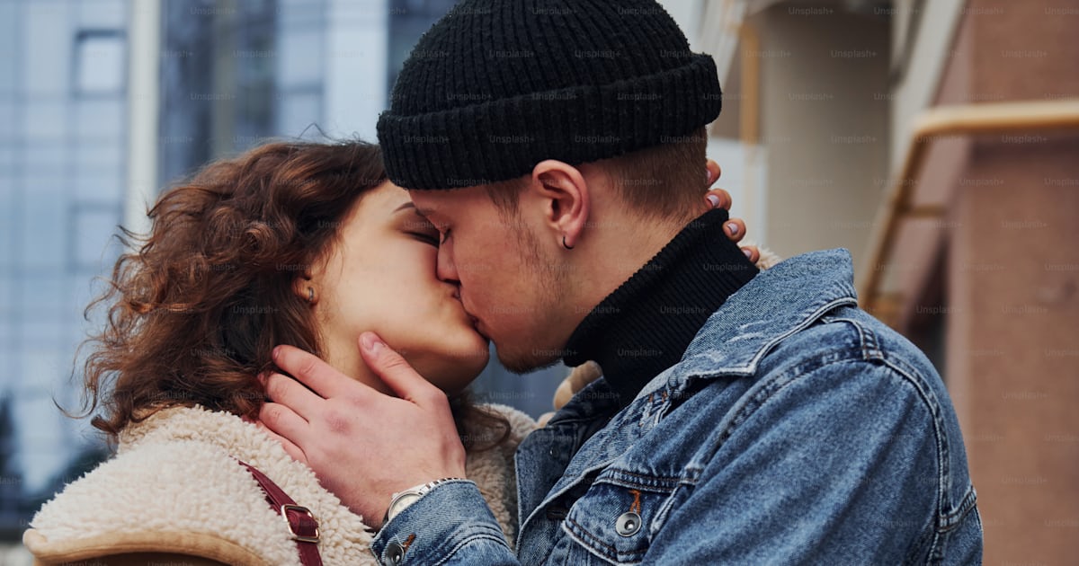 カジュアルな暖かい服を着た陽気なカップルが、ビジネスビルの近くの街で屋外でキスをしています。の写真 Unsplashの一体の画像