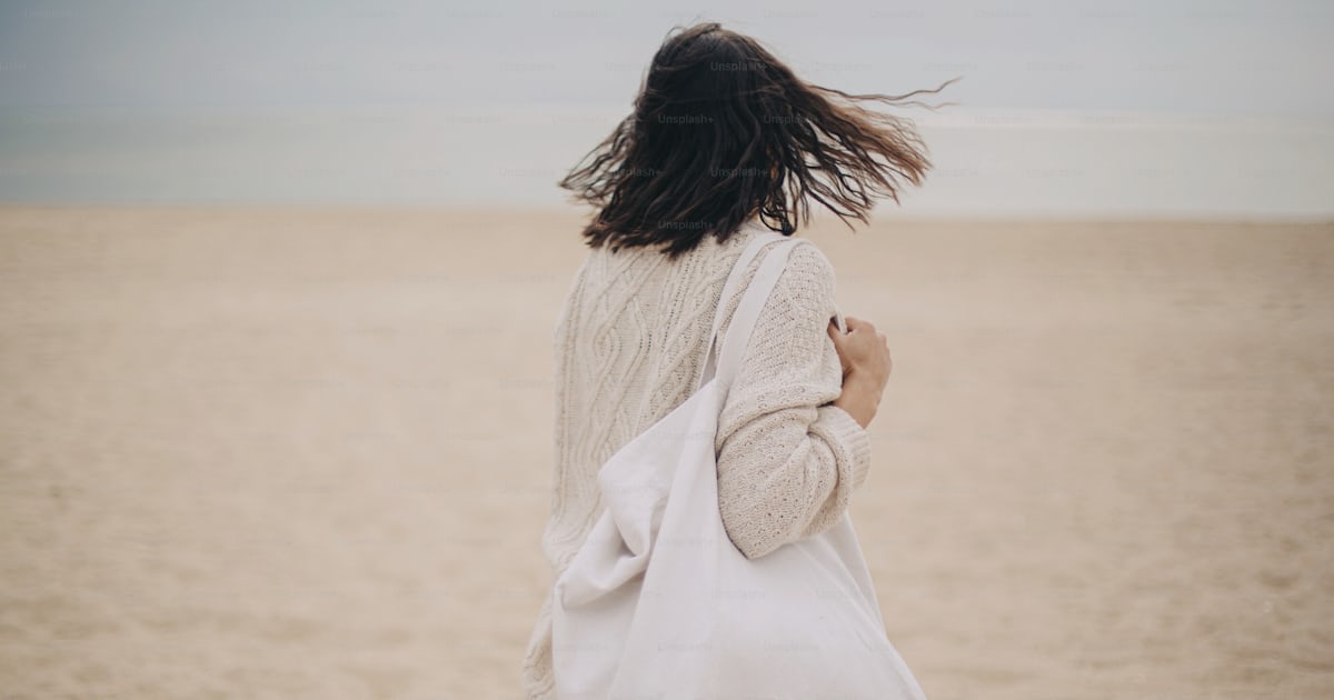 바람이 부는 머리와 토트백을 들고 모래 해변에서 바다로 걸어가는 아름다운 세련된 여성 평온한 순간 니트 스웨터를 입은 세련된