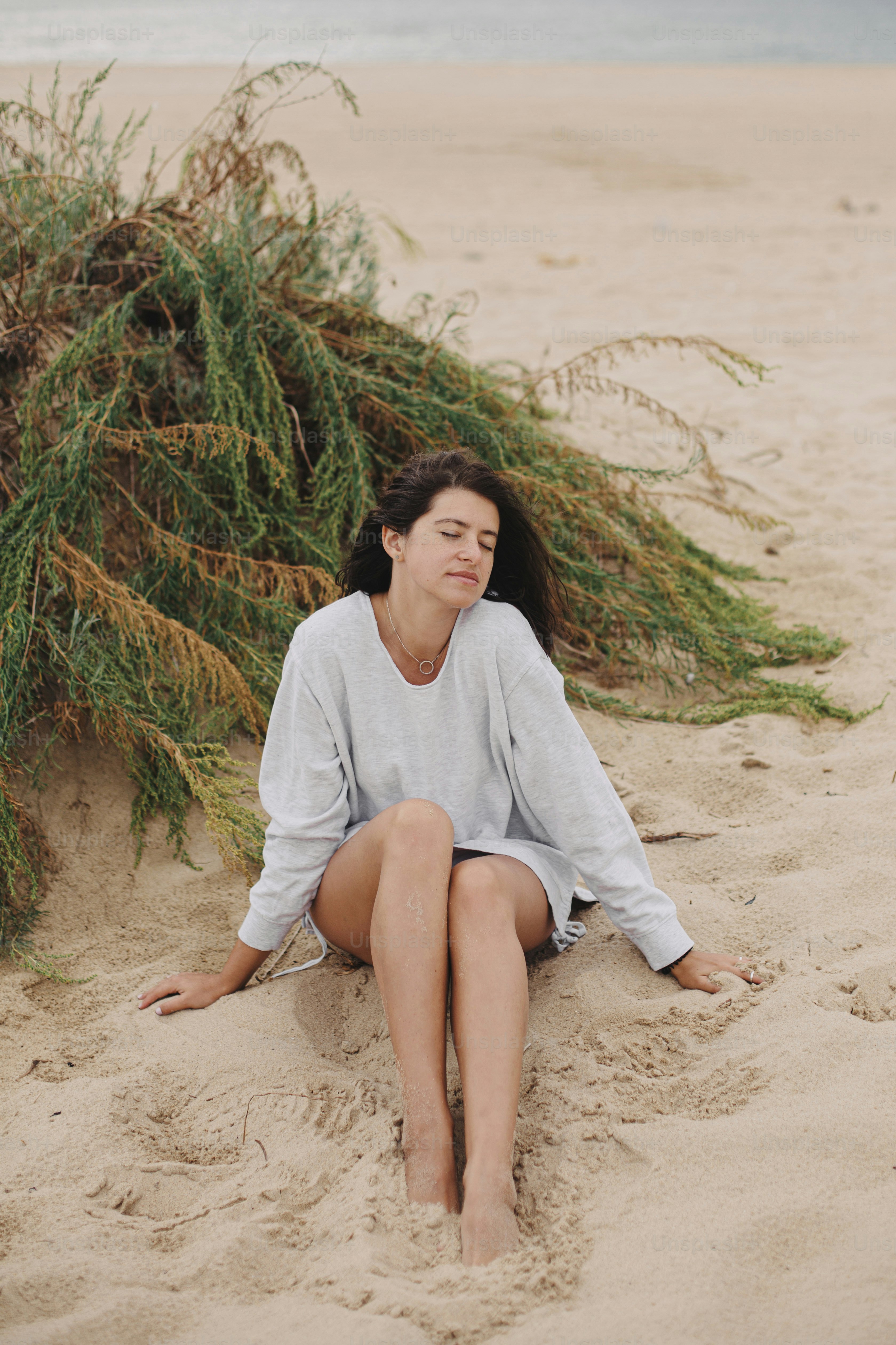 Carefree beautiful woman with windy curly hair sitting on sandy beach ...